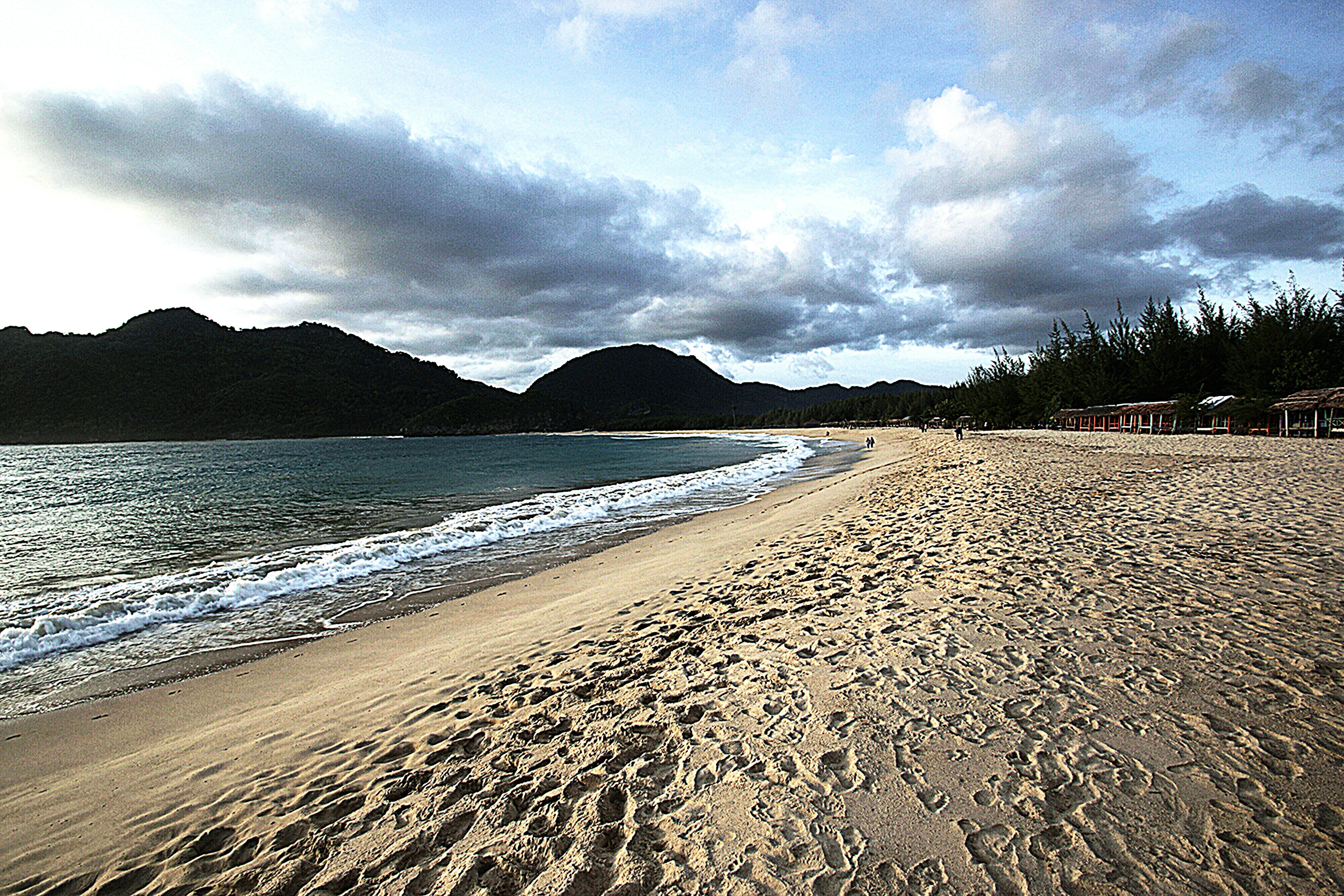 Sandy beach with footprints stretching toward gentle waves and distant hills under a partly cloudy sky.