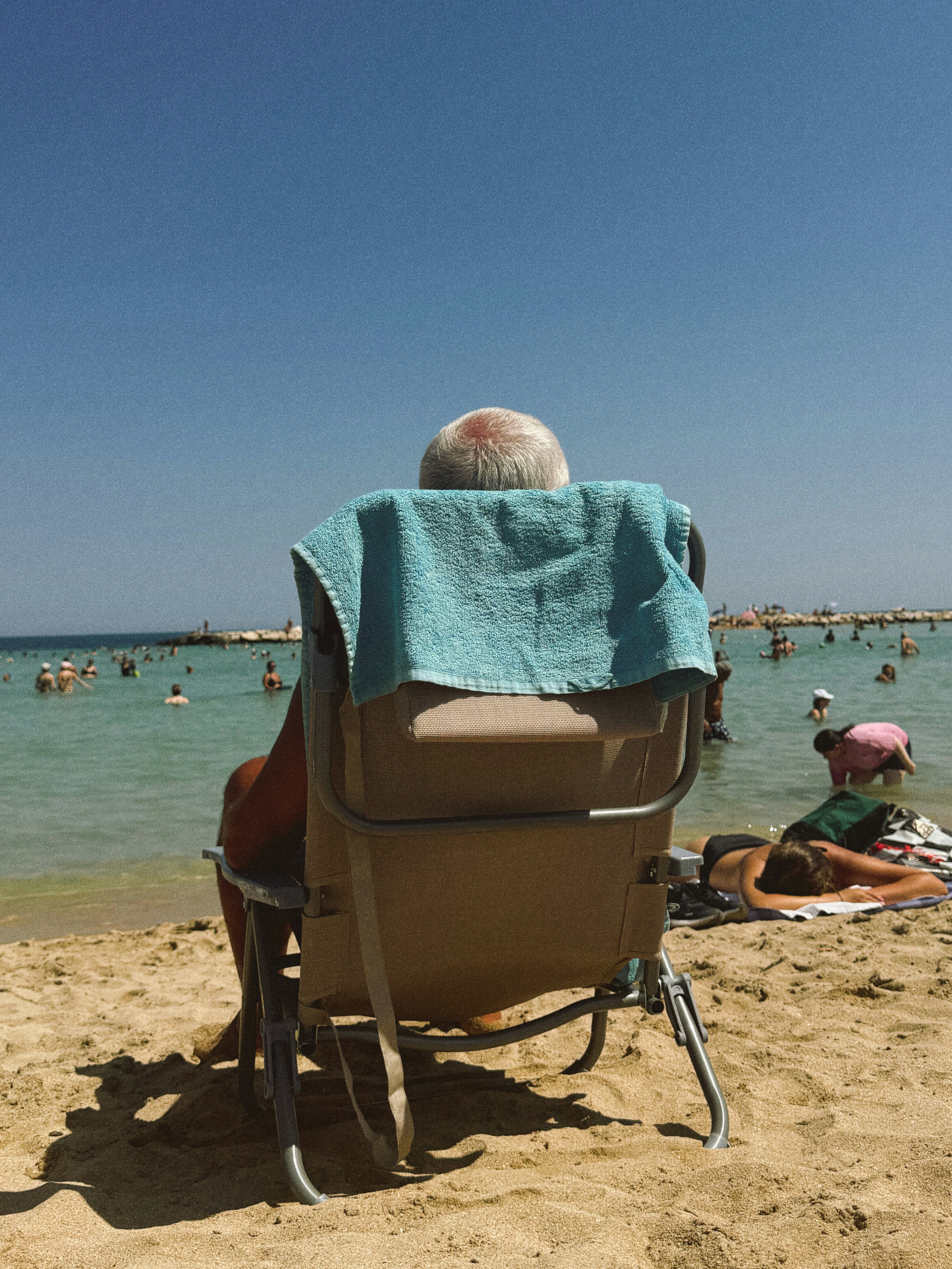 Sunbathers on a crowded beach with an elderly person reclining in a chair under a turquoise towel, facing the calm sea. This is a candid seaside scene capturing leisure and social activity at the water's edge.