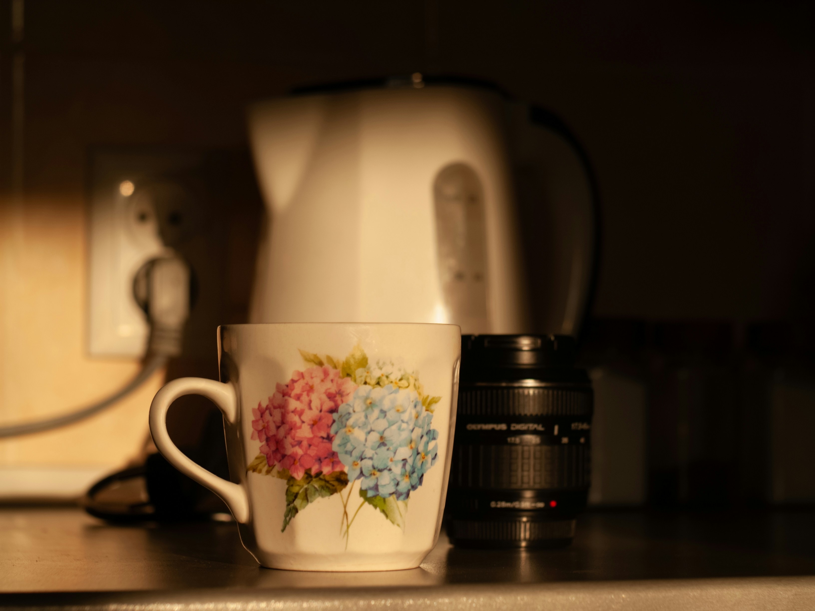 A ceramic mug with hydrangea decals sits on a kitchen counter beside a camera lens; a kettle and outlet are visible in the dim background.