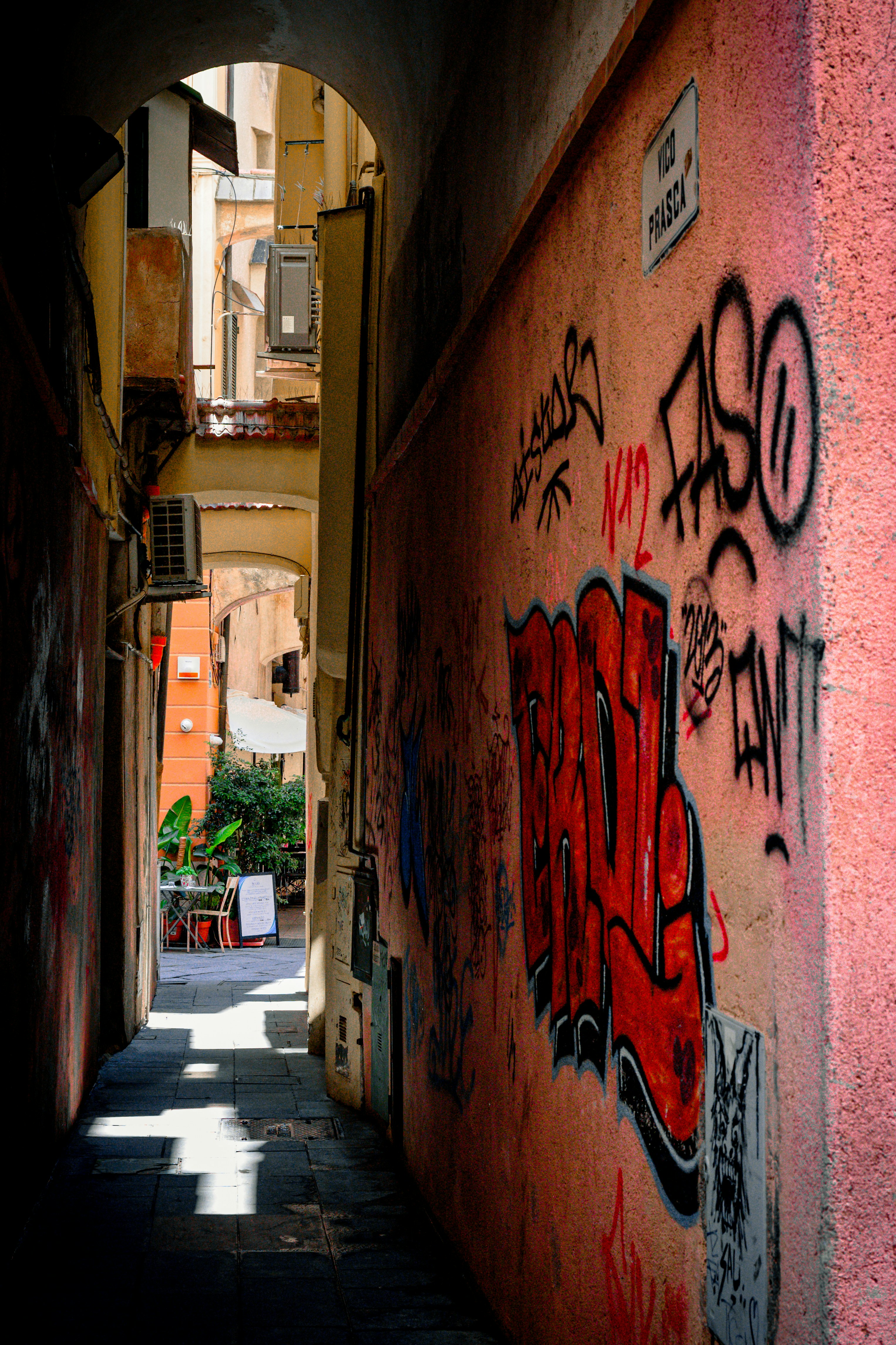 A narrow alleyway with pink graffiti-covered walls leading to a sunlit courtyard