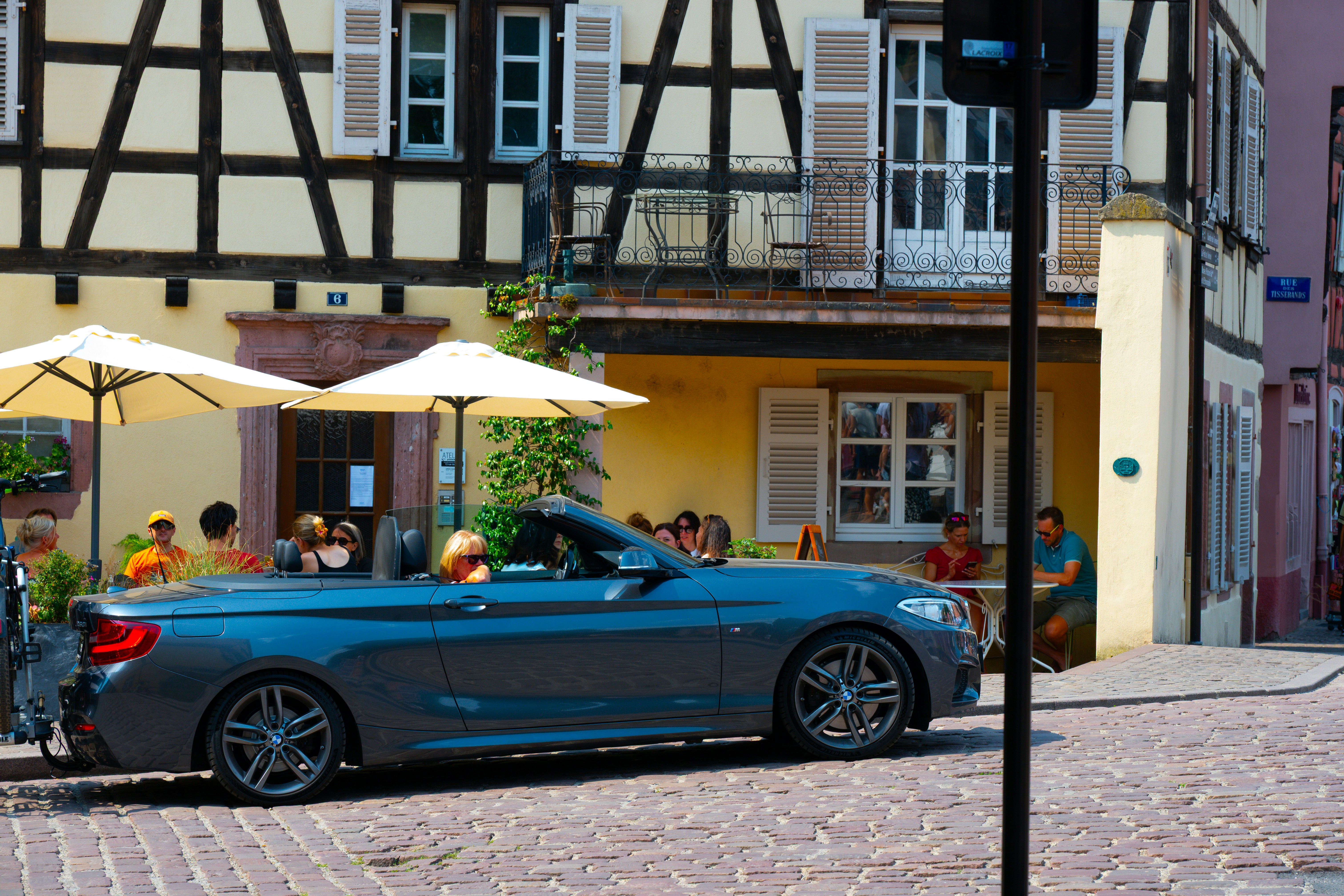 Sapphire-blue convertible parked along a cobblestone street beside a café with outdoor seating and half-timbered buildings.