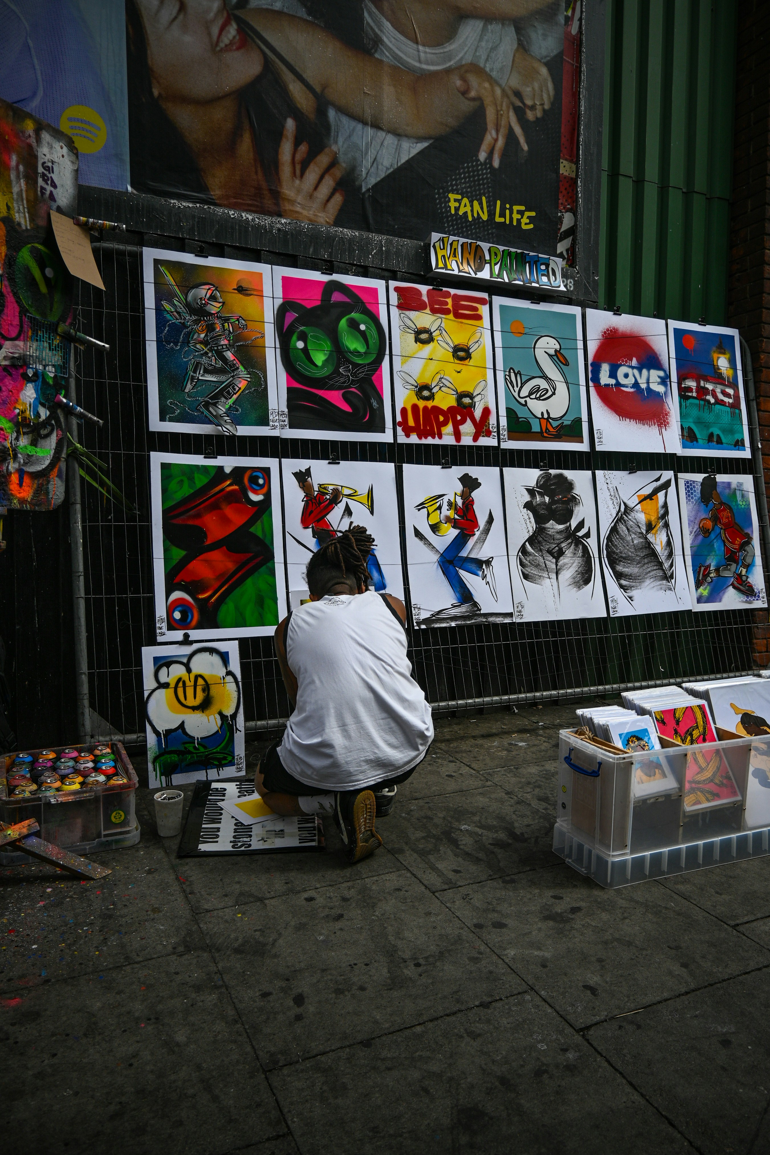 Colorful street-art prints displayed on a wall, with an artist crouched and painting below. A collection of bold, graphic illustrations fills the foreground while a large poster looms above.