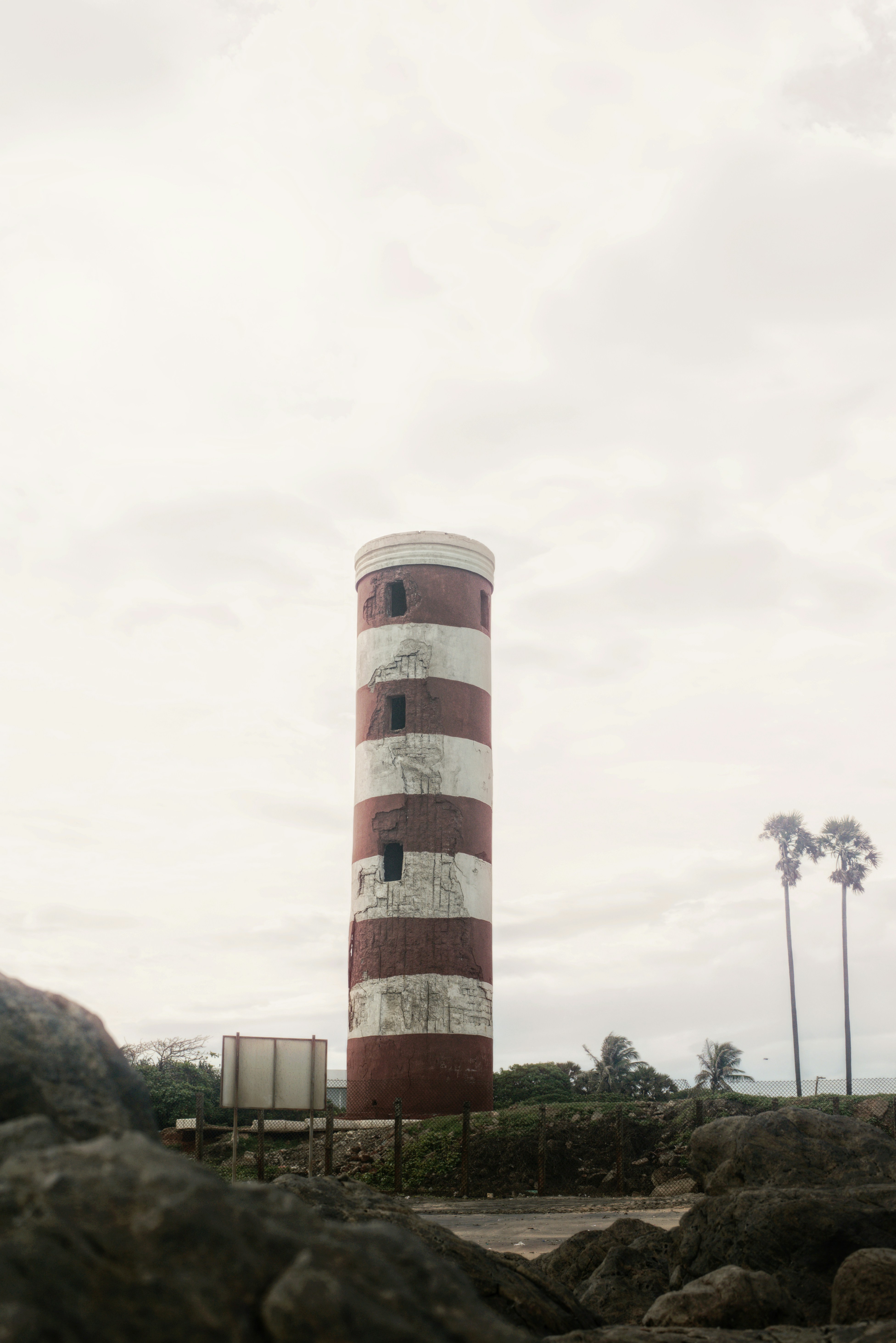 A weathered striped lighthouse rises above a rocky foreground with distant palm trees under a soft, pale sky. A quiet coastal scene highlighting weathered architecture and tranquil mood.