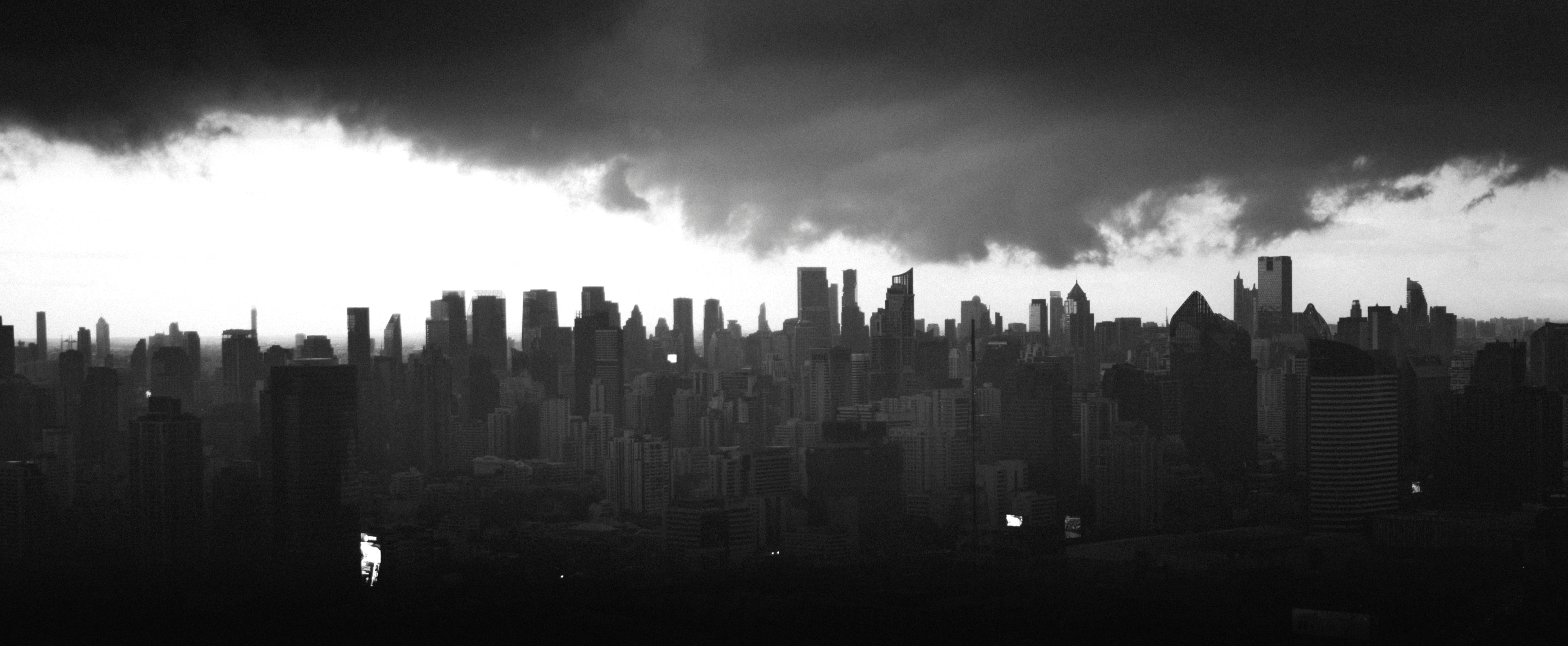 Dark urban skyline silhouettes beneath a looming storm, with a dense cloud deck pressing low over the horizon.