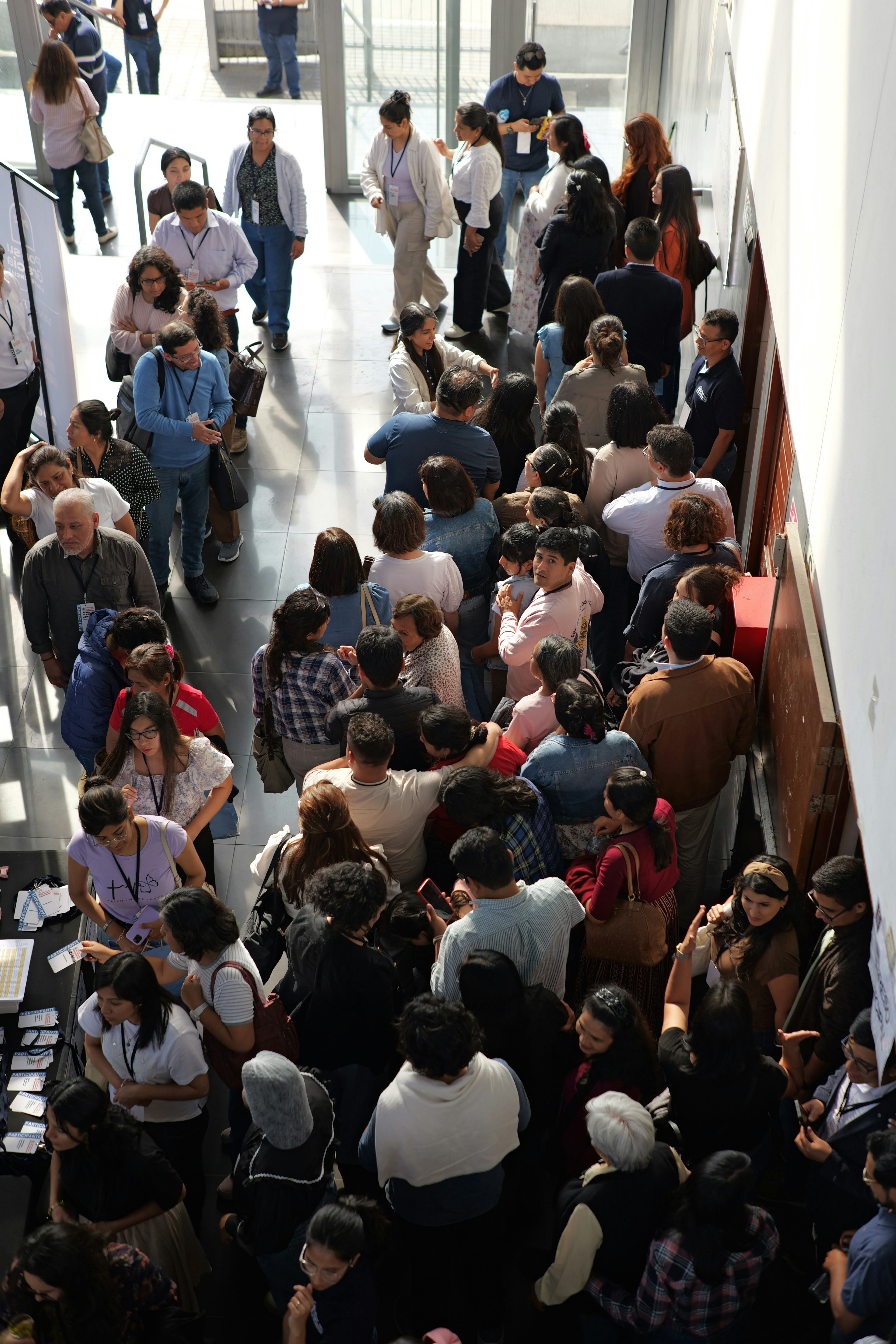 Dense crowd gathered in a bright, modern hallway; attendees mingle, walk, and consult displays along the sides in a conference or exhibition space.