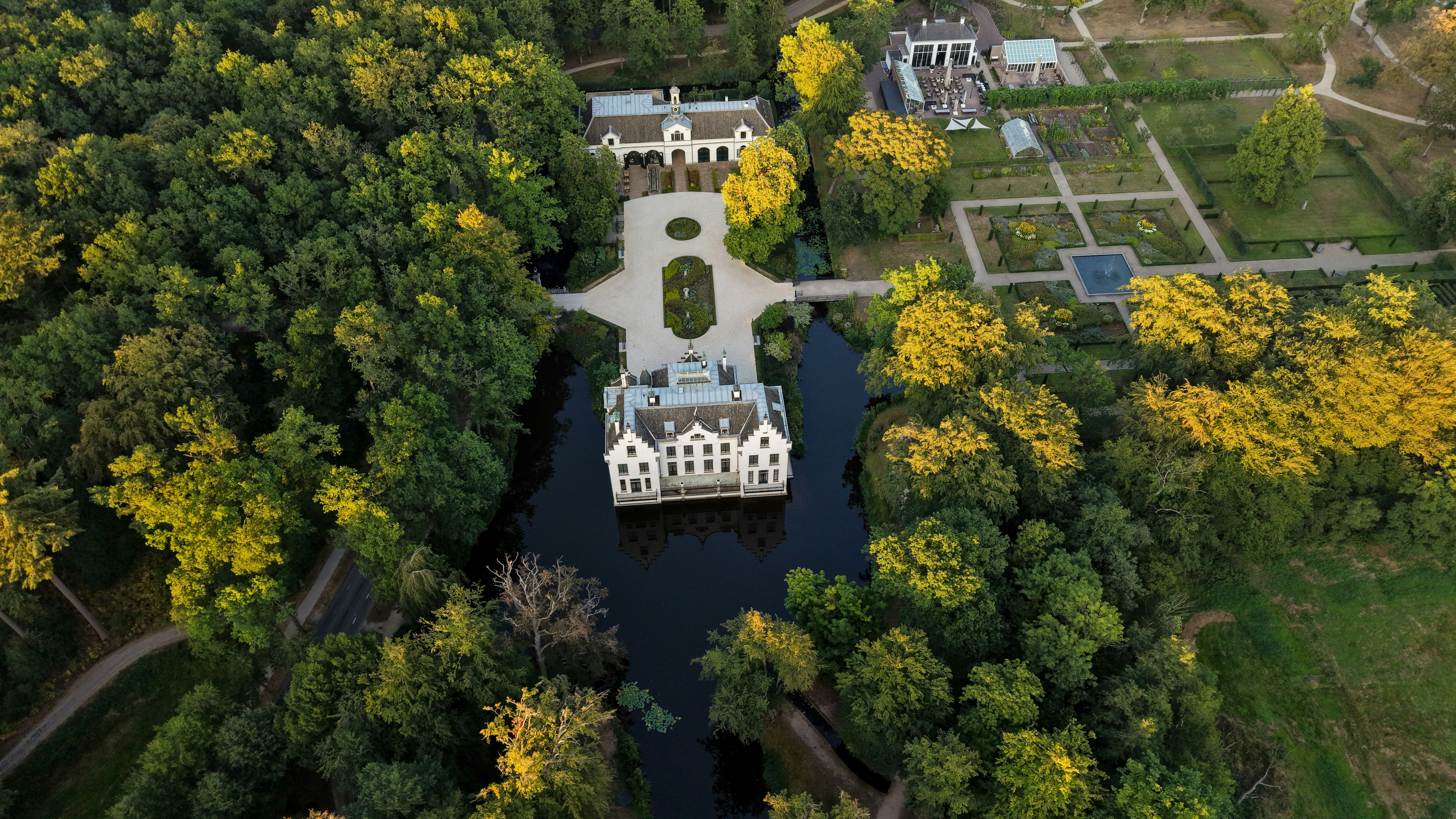 A grand manor sits centered on a reflective pond, encircled by a formal garden and dense autumn trees, viewed from above in golden-hour light.