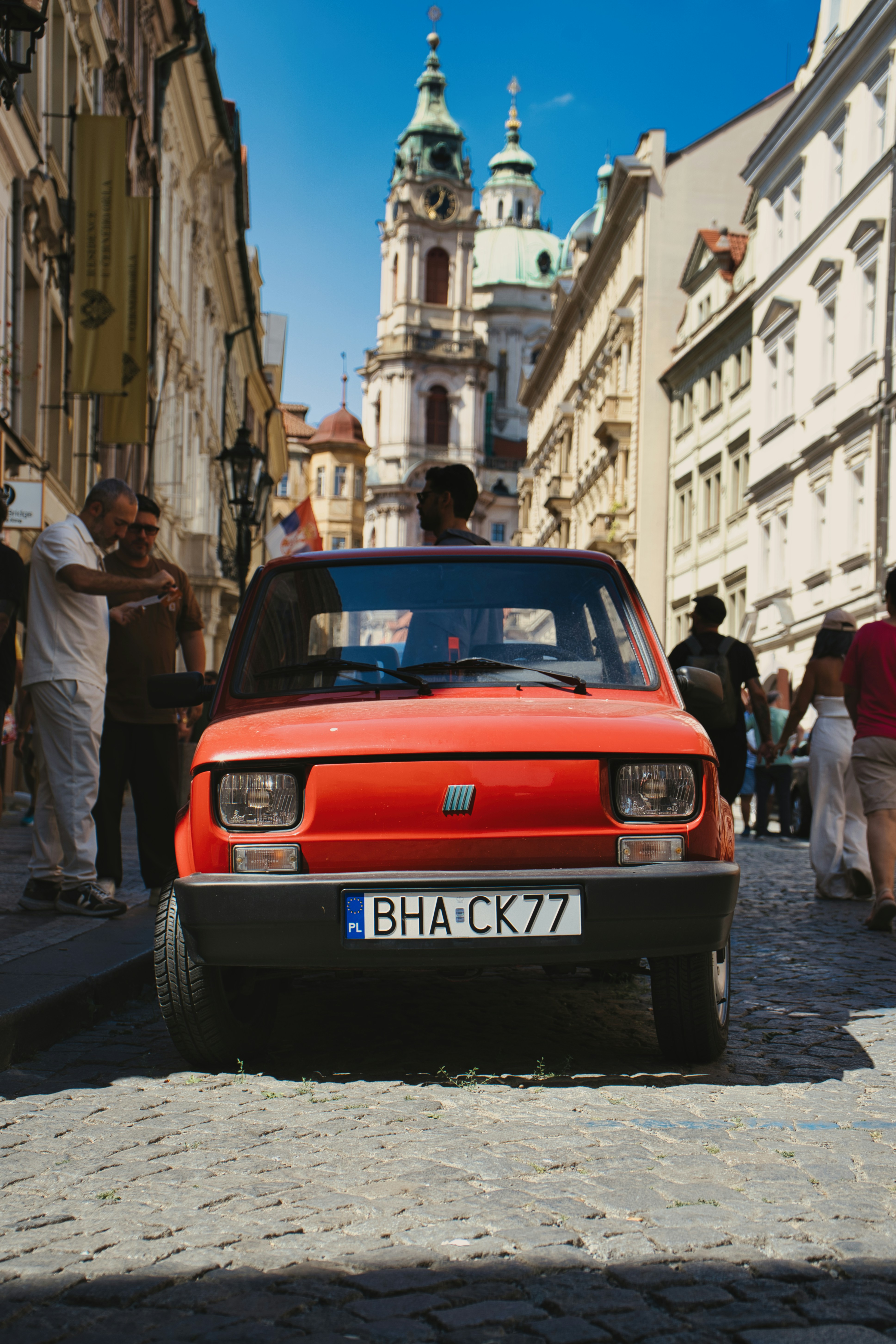 A vibrant red vintage Fiat 126p parked on a cobblestone street in a European Old Town, framed by classic architecture and a historic church tower in the background under a sunny sky.