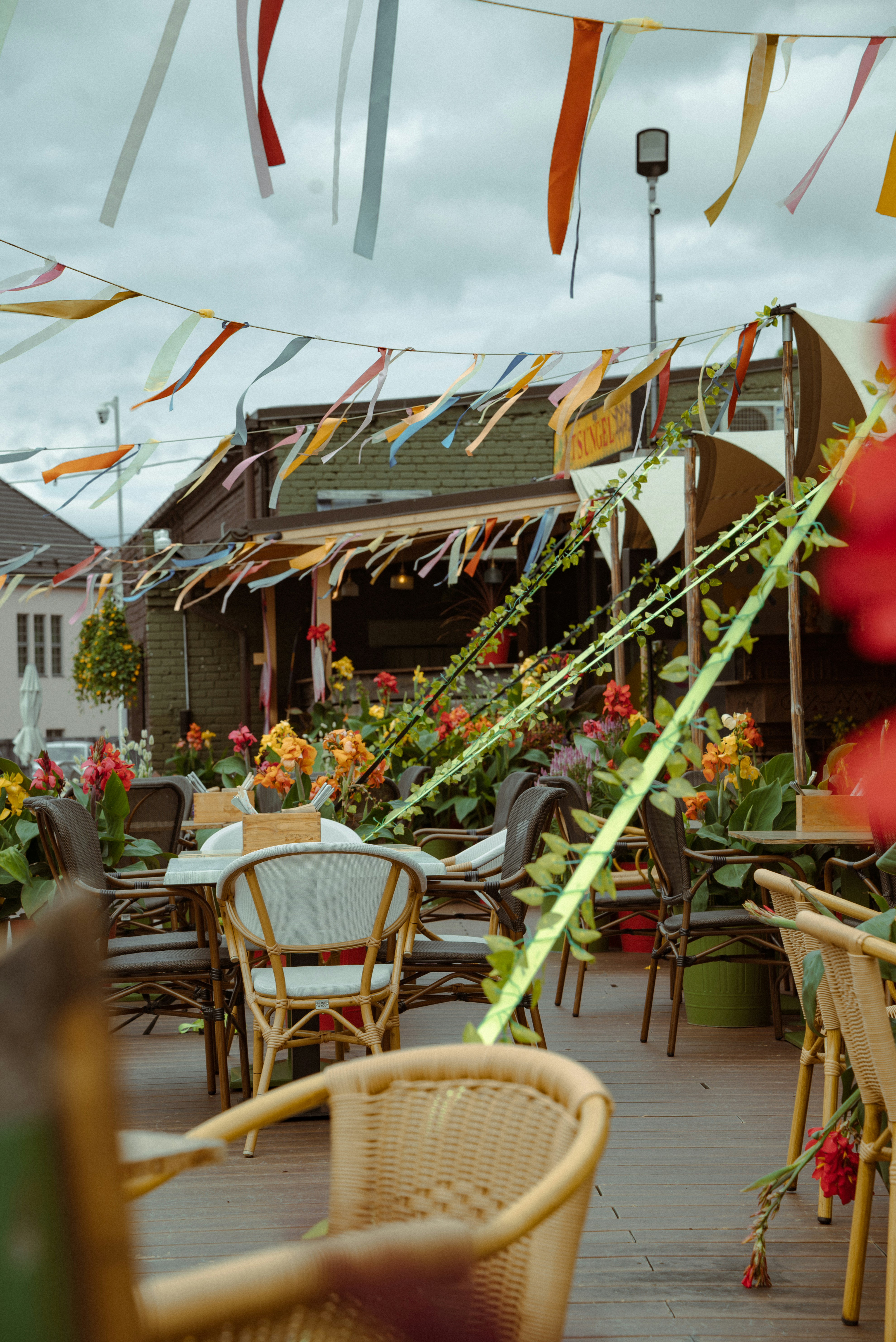 A sunlit outdoor café adorned with colorful bunting and blooming plants, inviting diners over wicker chairs and wooden tables.