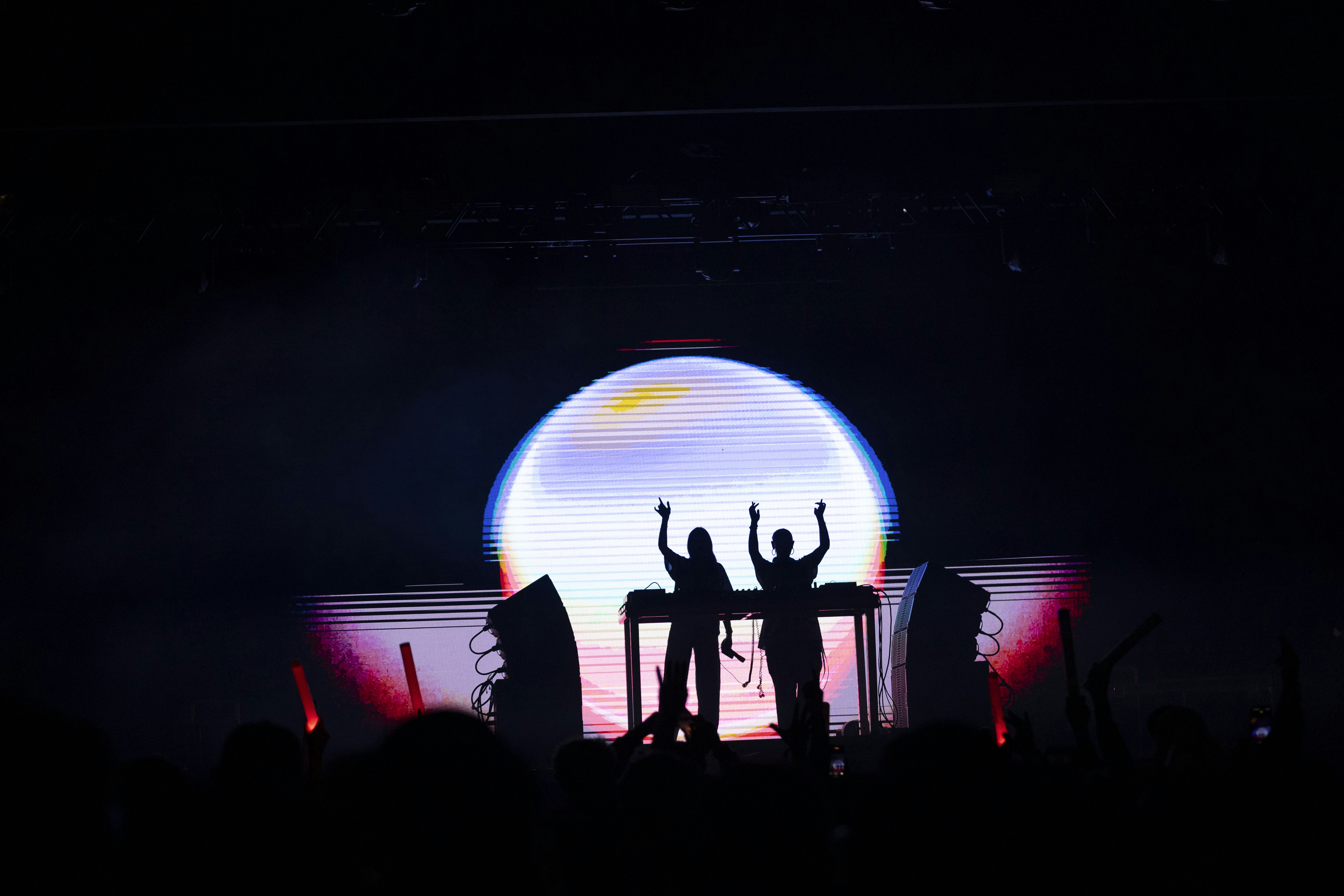 Silhouetted performers at a live show stand in front of a circular neon light, with stage lighting and a dark audience in the foreground. This is a concert scene with a bold glow behind the subjects.