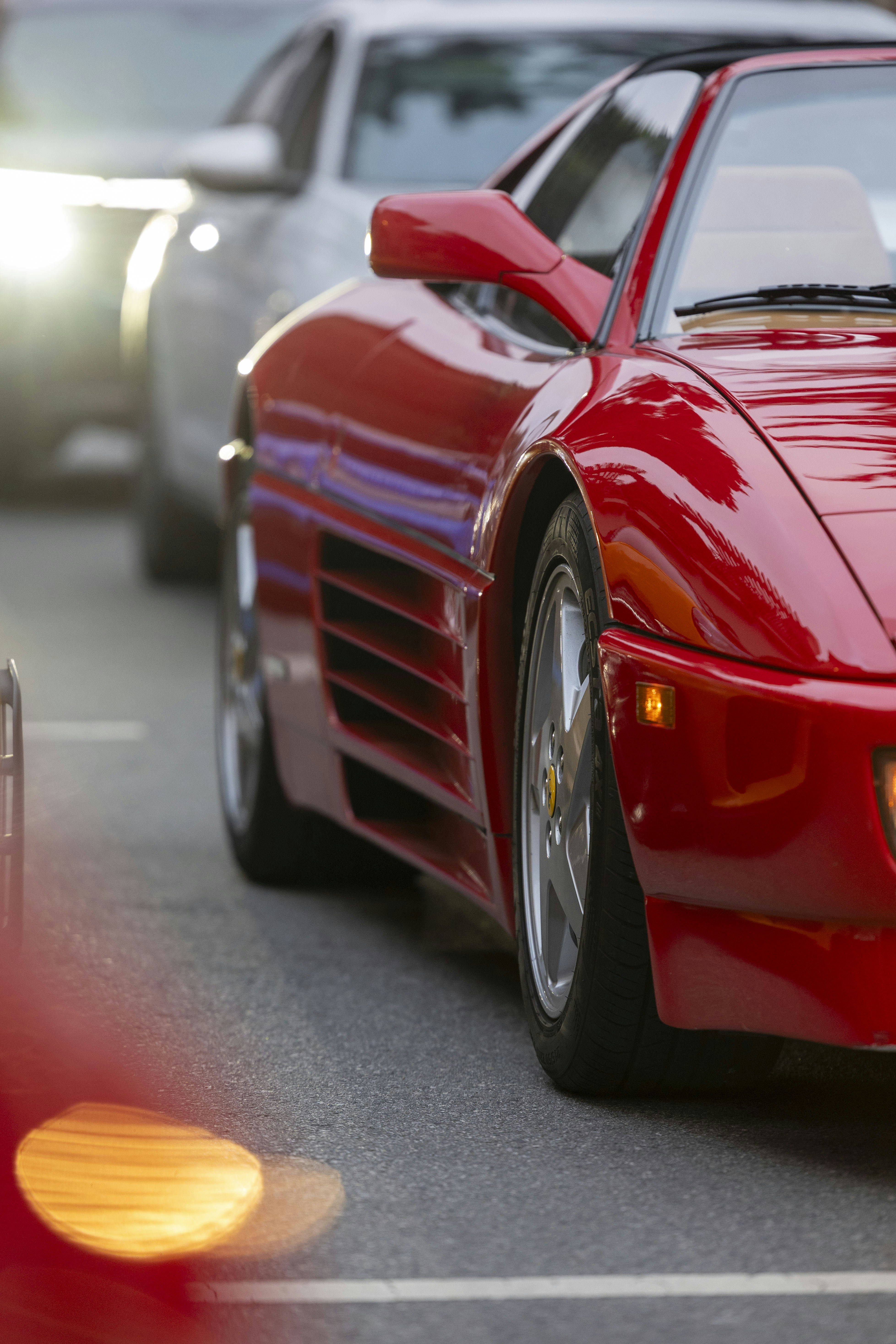 Crimson sports car lines the frame, with a sharp focus on the front quarter and wheel detail against a blurred background; a hint of another vehicle behind suggests a street scene. This is a high-contrast automotive shot.