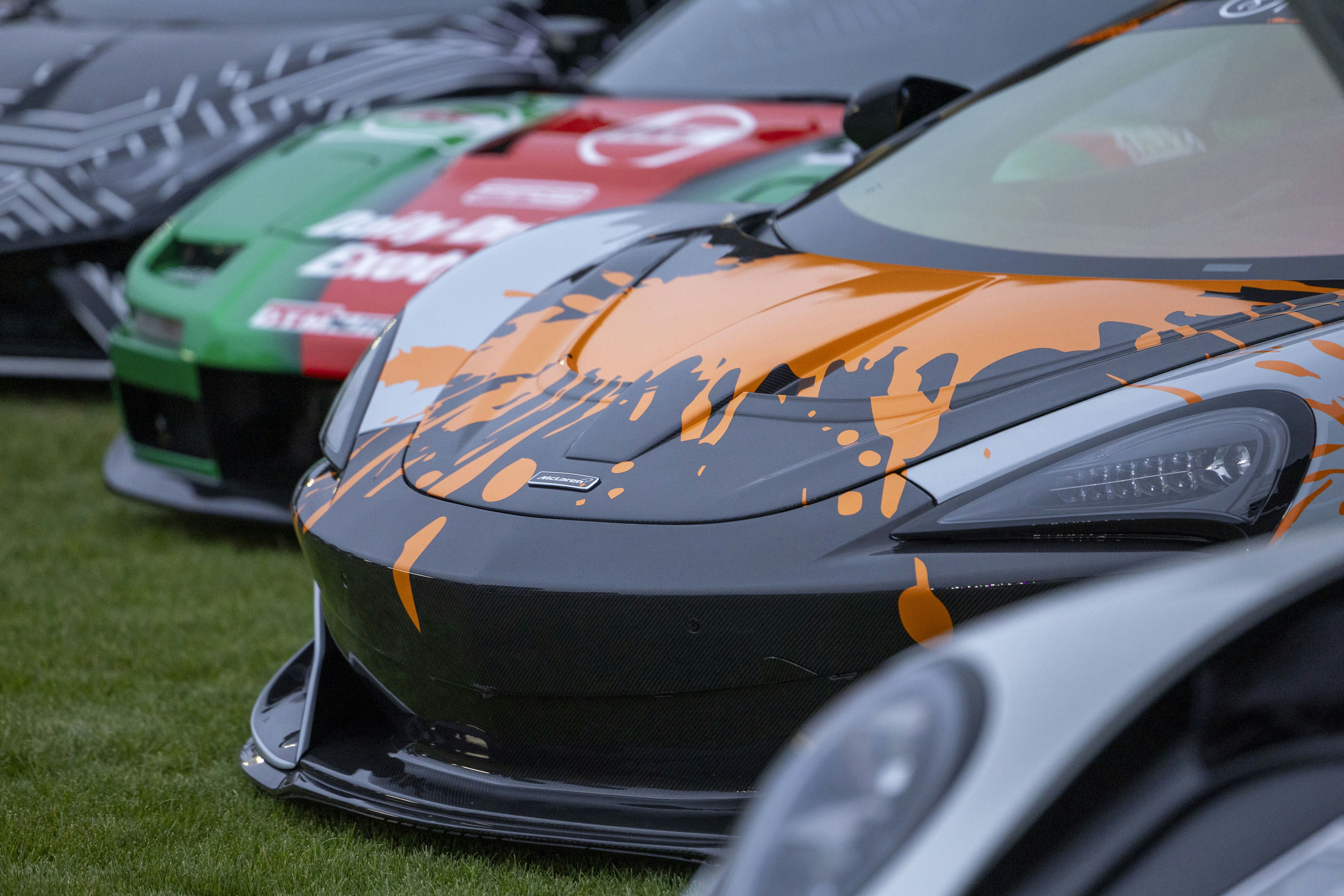 Glossy orange-splattered racing car in the foreground with other colorful cars blurred in the background, parked on grass at a show.