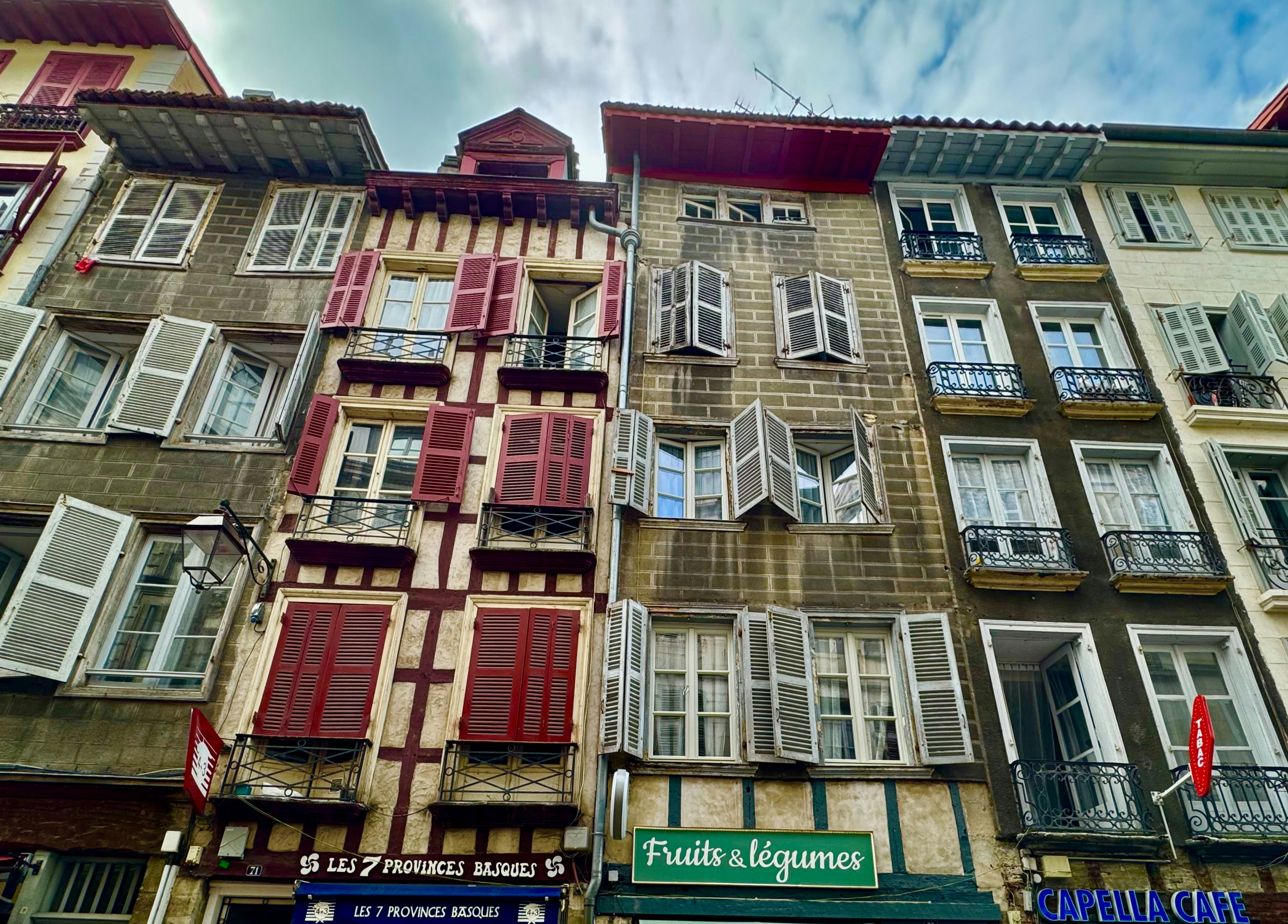 A row of vibrant, shuttered façades lines a European street, with fruit and cafe signs beneath. The architecture features varied window styles and decorative timber framing, capturing a lively urban storefront scene.