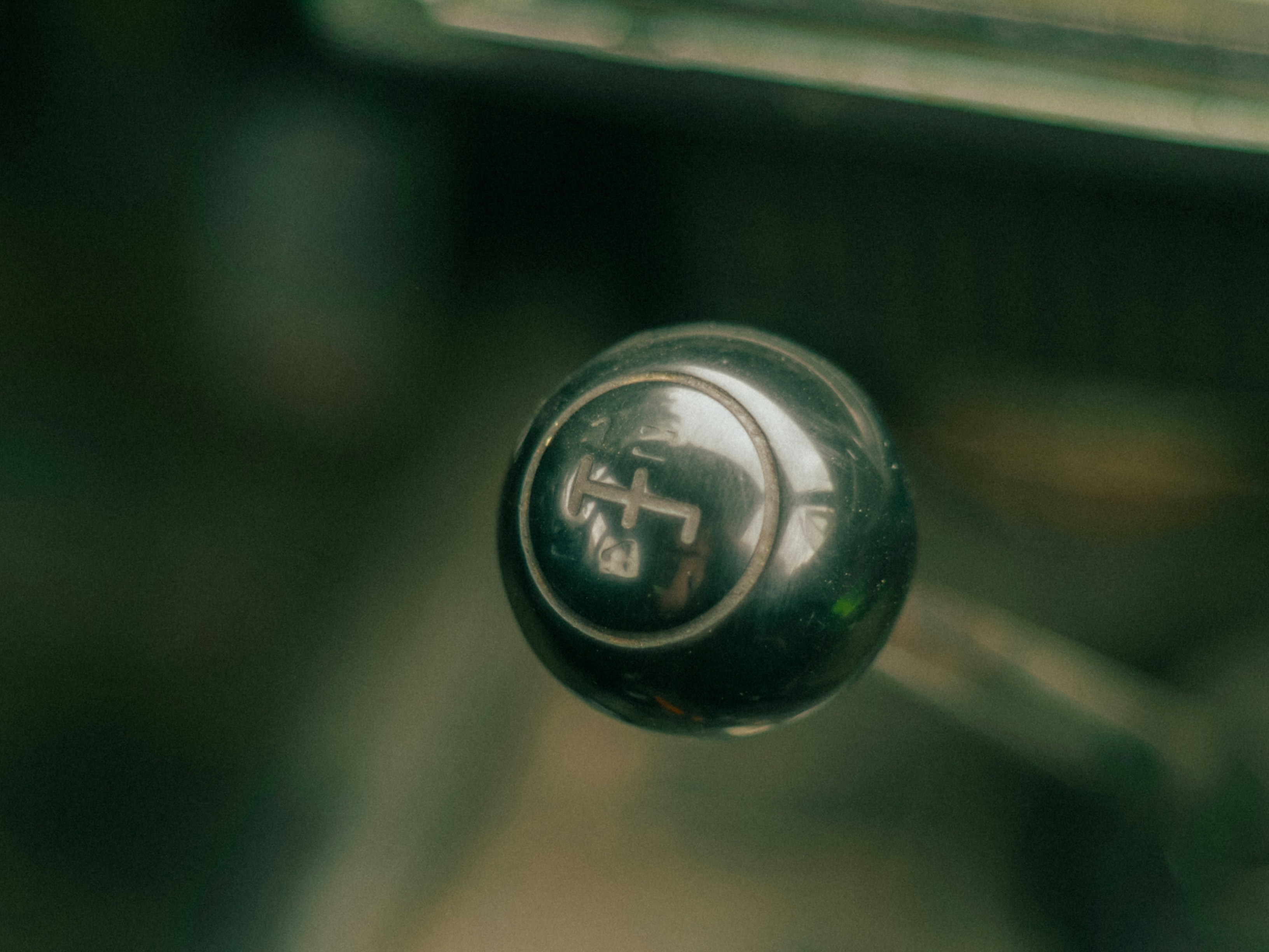 Close-up of a round gear knob with a metallic surface, slightly worn, set against a shallow bokeh-green background.