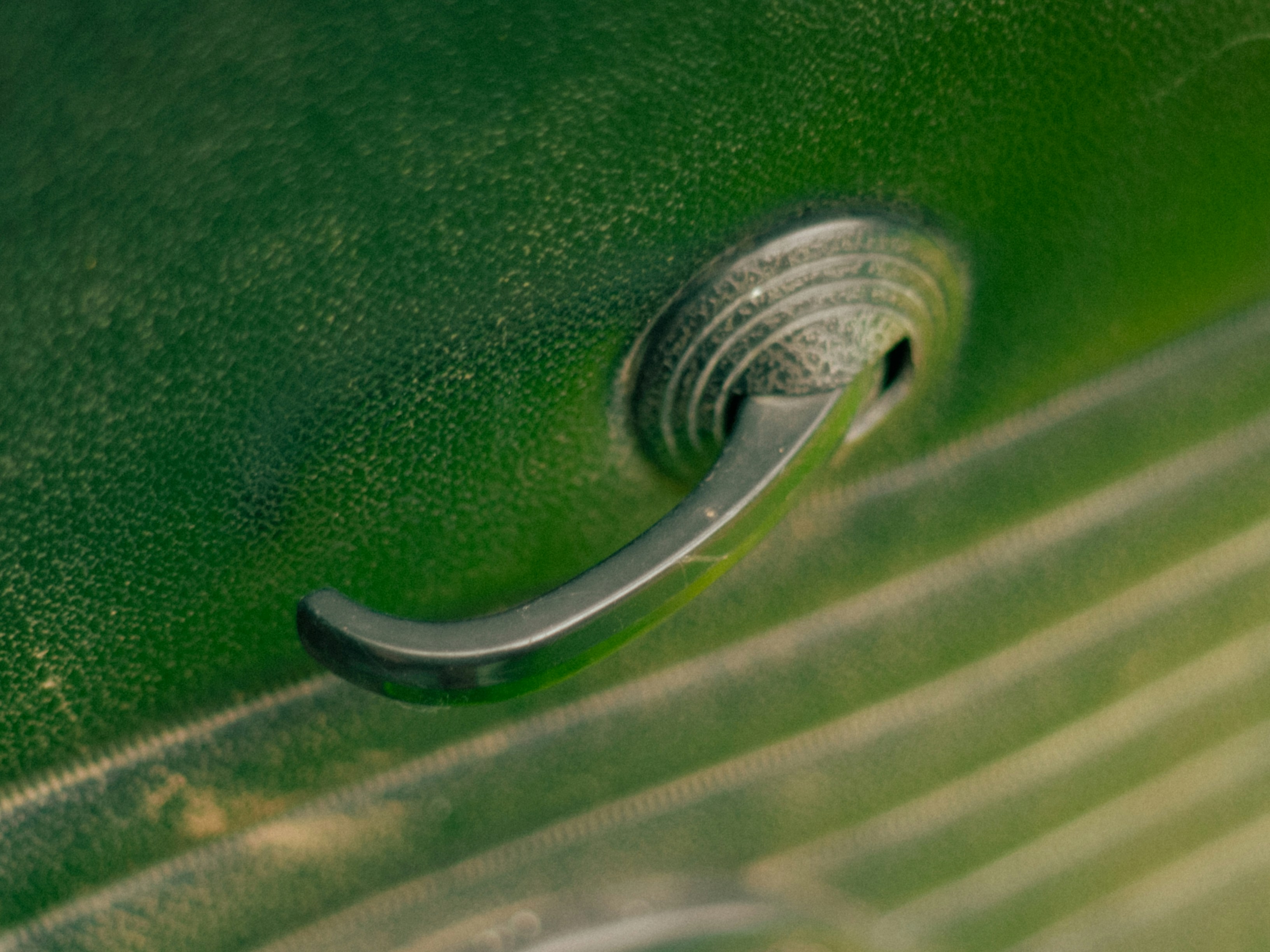 Close-up of a metallic door handle set into a textured green surface, with subtle aging and embossed circular escutcheon. The composition emphasizes the curved lever and the surrounding material texture.