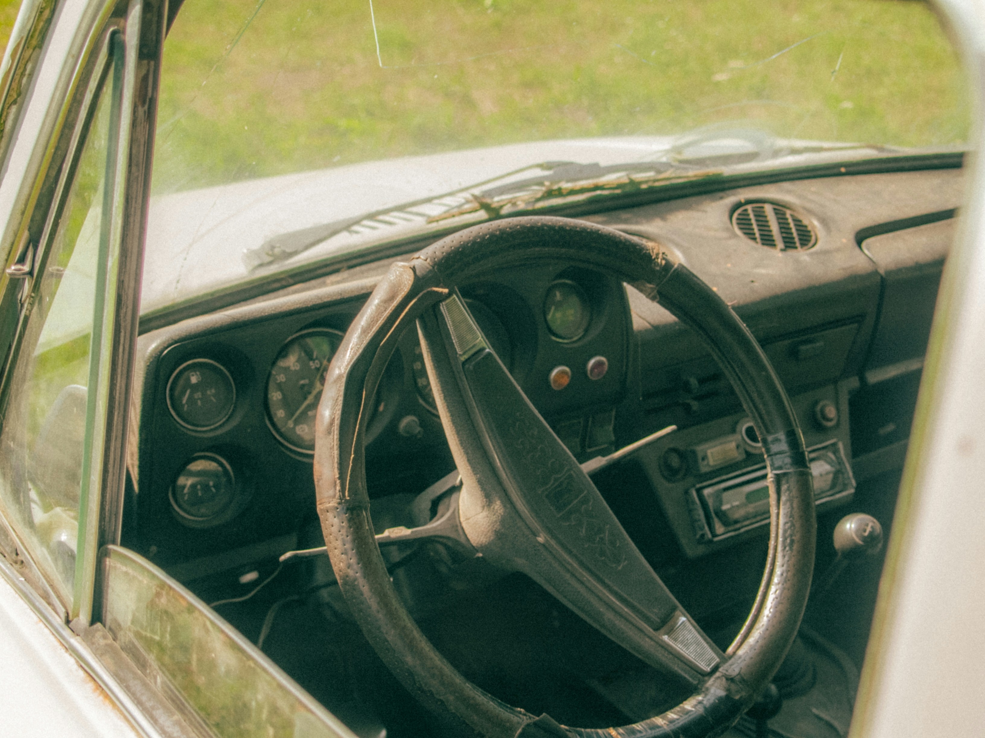 Close-up view into a classic car's cockpit, highlighting the worn steering wheel, dashboard gauges, and retro controls. The scene emphasizes the vehicle's aged character and timeless interior.
