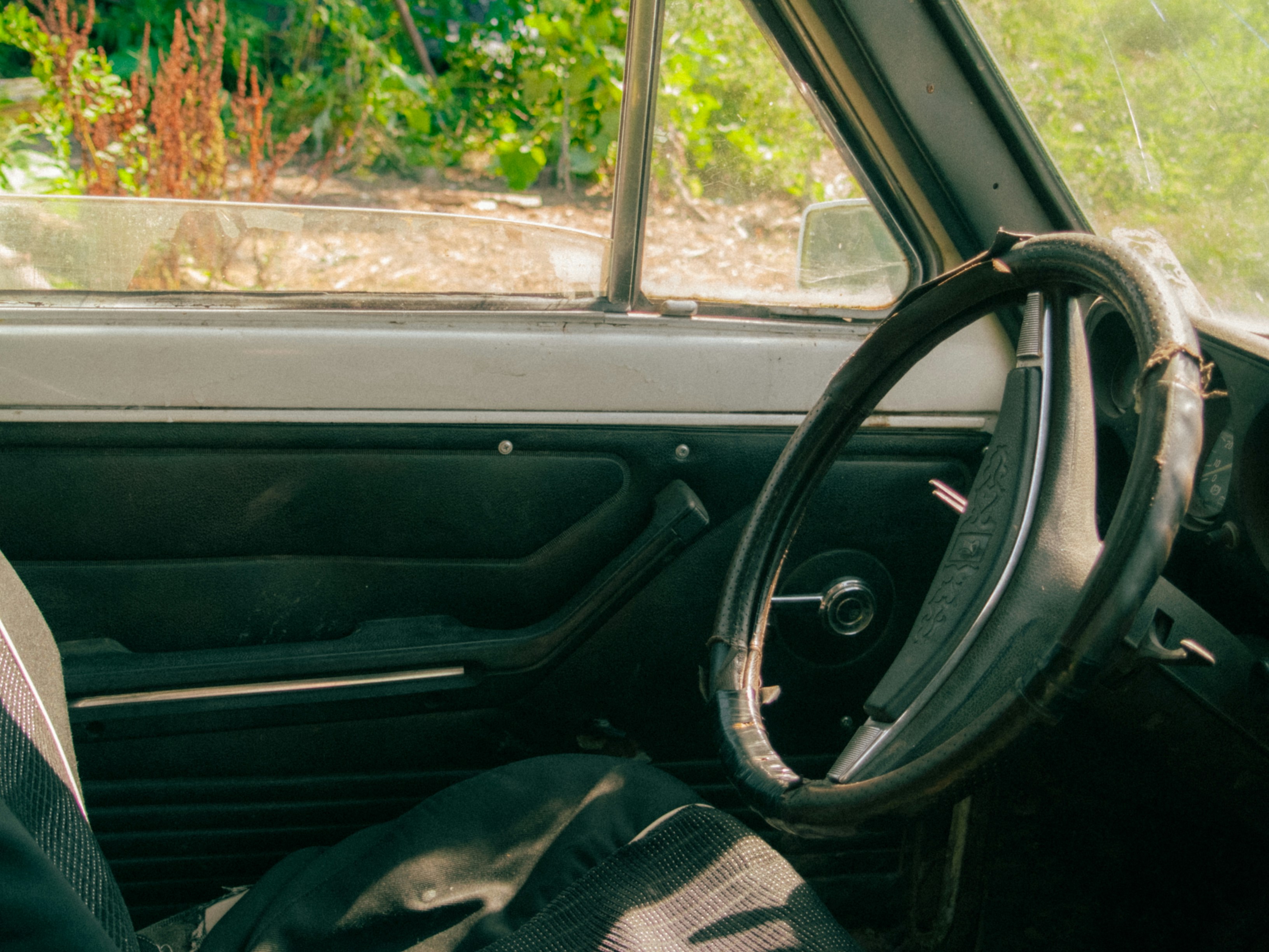 A vintage car interior with a weathered steering wheel and slightly worn upholstery, illuminated by natural daylight from the open window. The scene focuses on the driver's area with a hint of exterior greenery visible through the window.