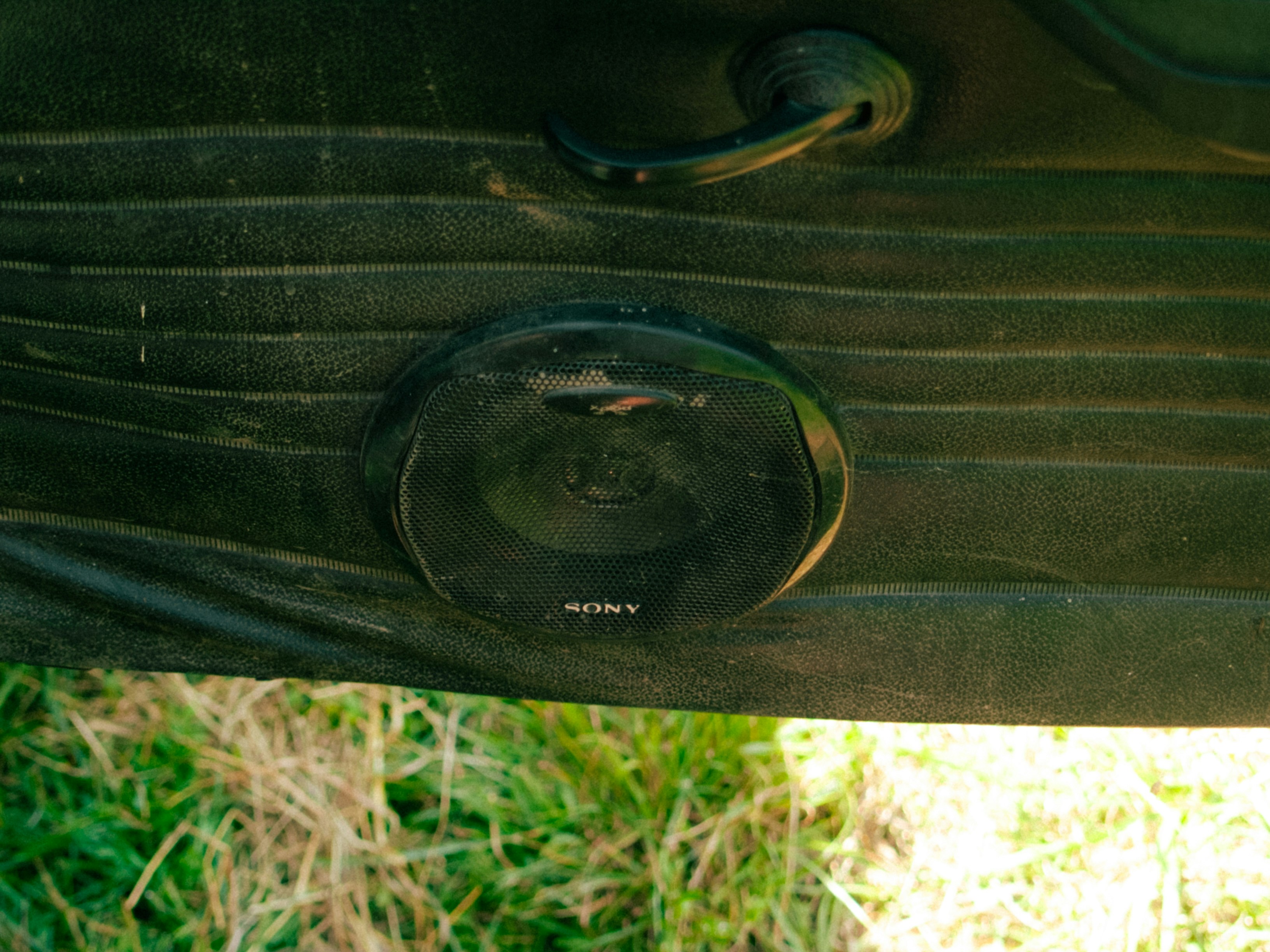 Close-up of a worn vehicle bed with a Sony speaker cap partially embedded in the bed liner, grasses visible underneath. The scene emphasizes texture and weathering.