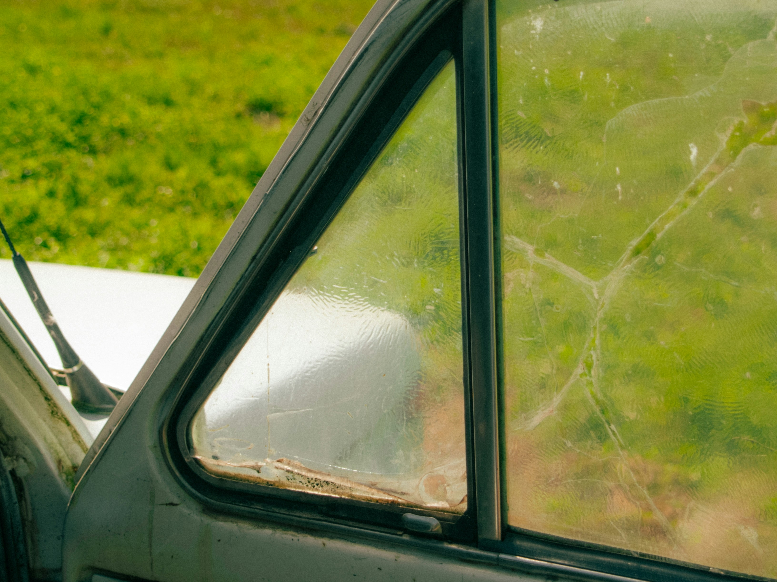 A close-up of a vintage car door and side window with a weathered frame, revealing a blurred green field outside and dusty glass; hints of a quiet, rural setting.