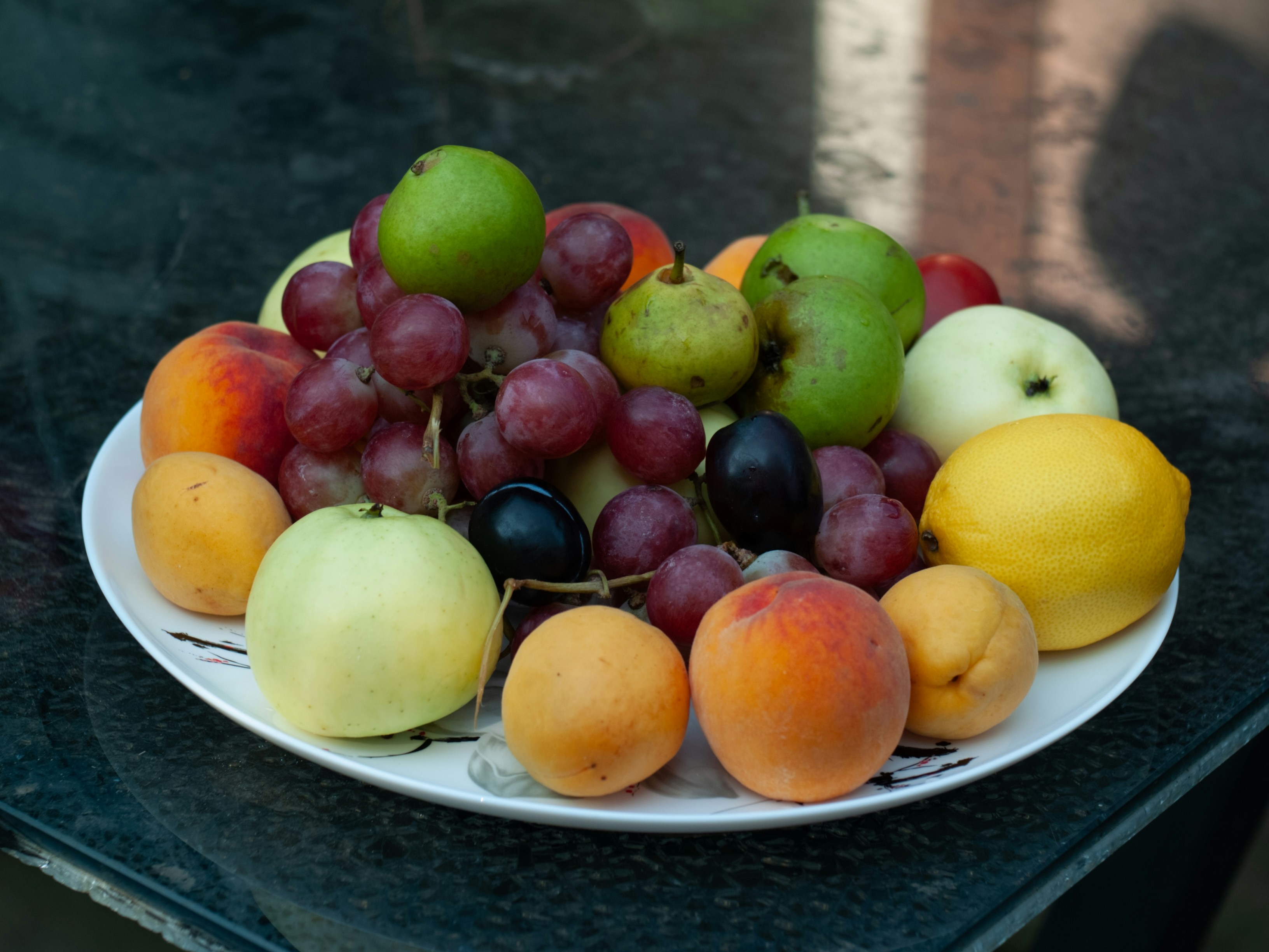 Colorful assortment of peaches, pears, grapes, and plums arranged on a white plate, showcasing natural fruit textures and a dark tabletop setting.