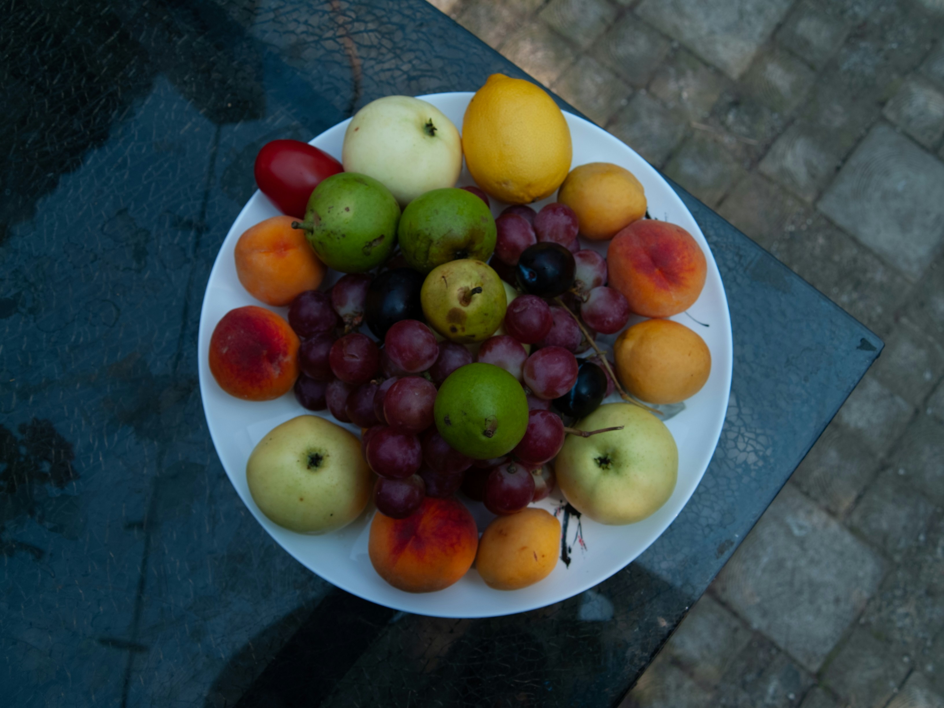 A white bowl brimming with apples, grapes, plums, and peaches sits on a reflective glass surface, framed by a stone-paved patio. The arrangement emphasizes vibrant colors and composition rather than a specific subject.