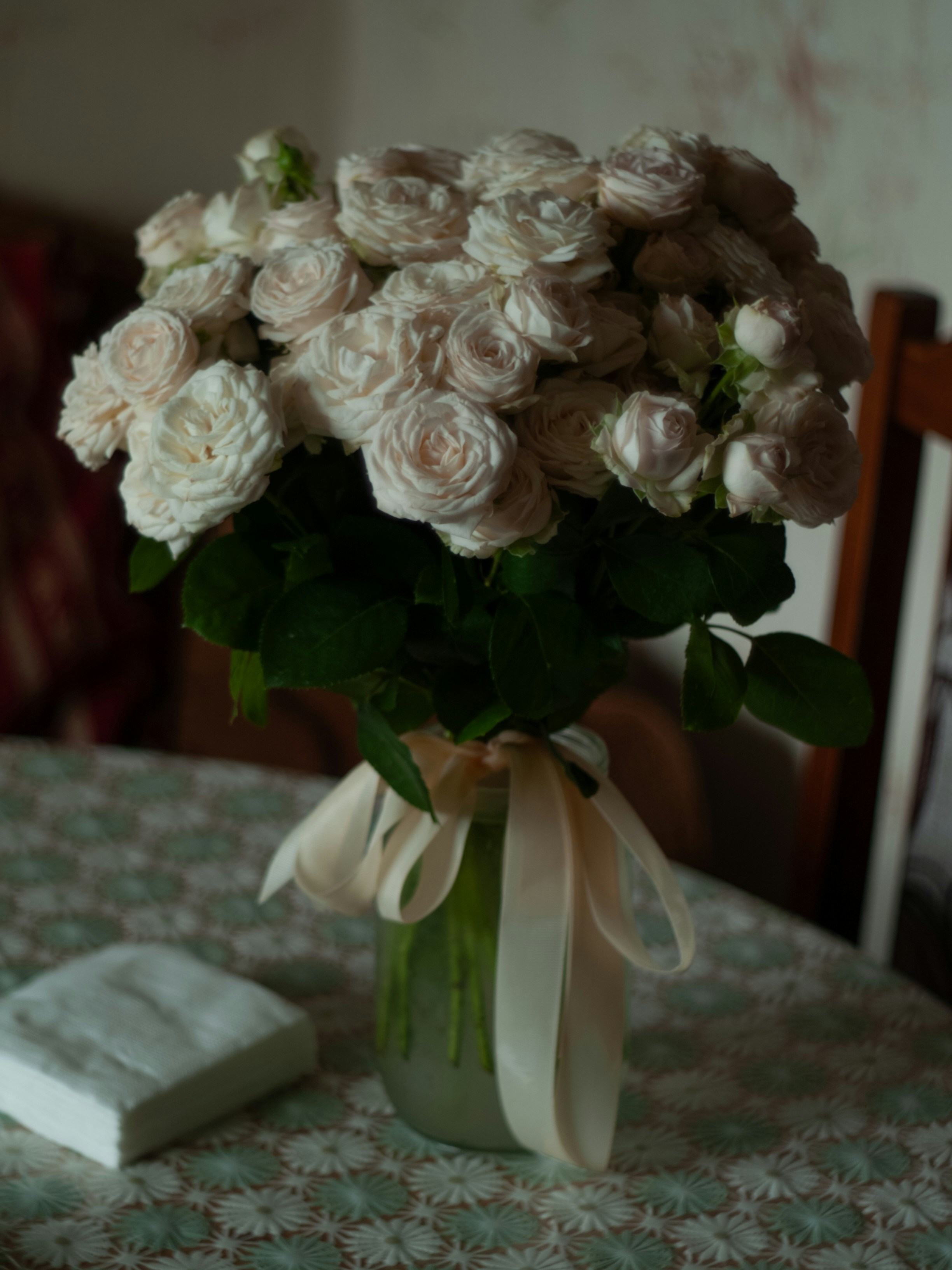 A lush bouquet of cream roses in a glass vase tied with a pale ribbon sits on a patterned tablecloth, subtle natural light illuminating the blooms.