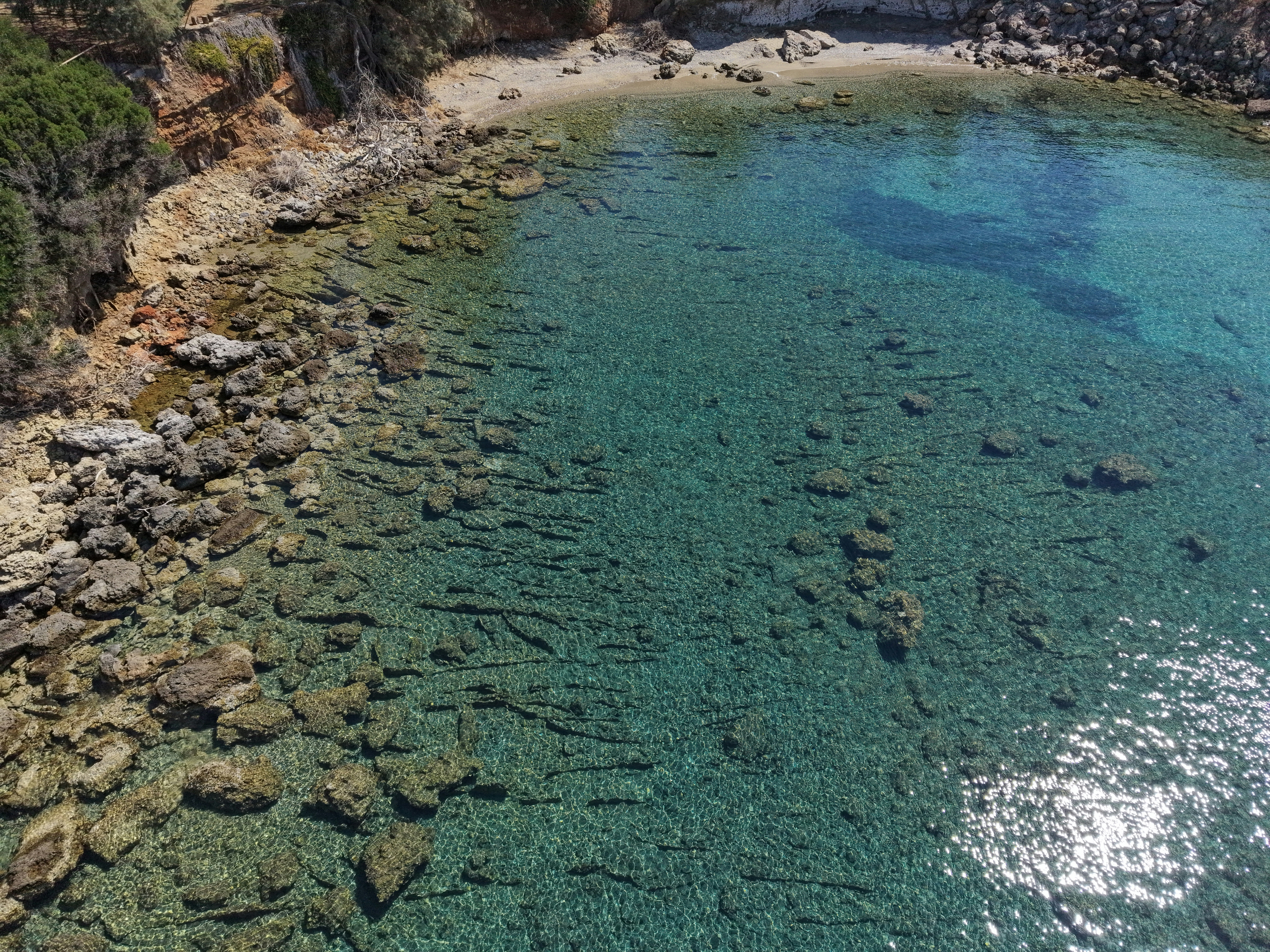 Crystal-clear turquoise water along a rocky coastline with sunlit shallows and scattered seaweed, revealing a shallow seabed and submerged rocks.