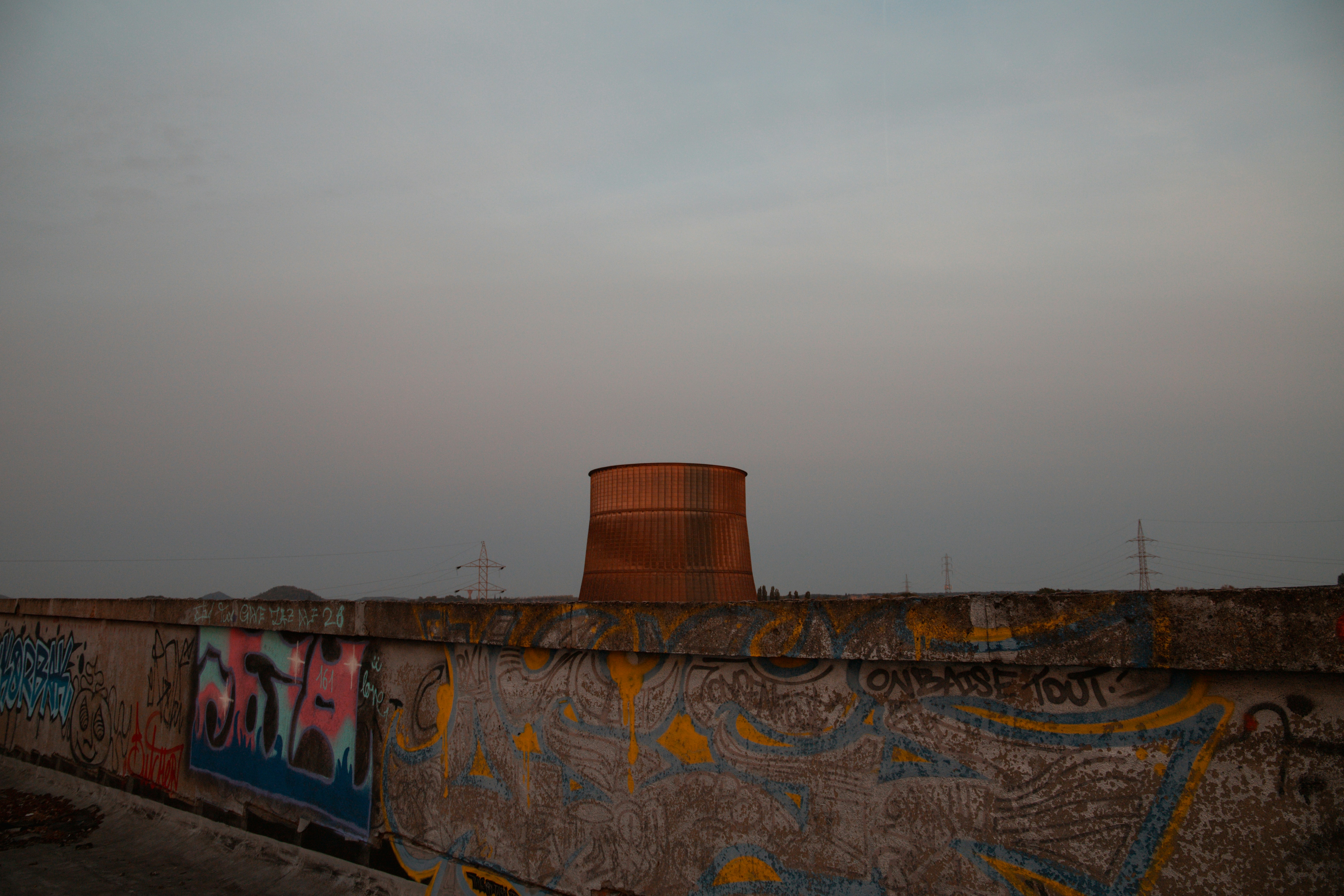 Rusted cylindrical vent rises above a graffiti-covered barrier, beneath a soft, dusky sky.