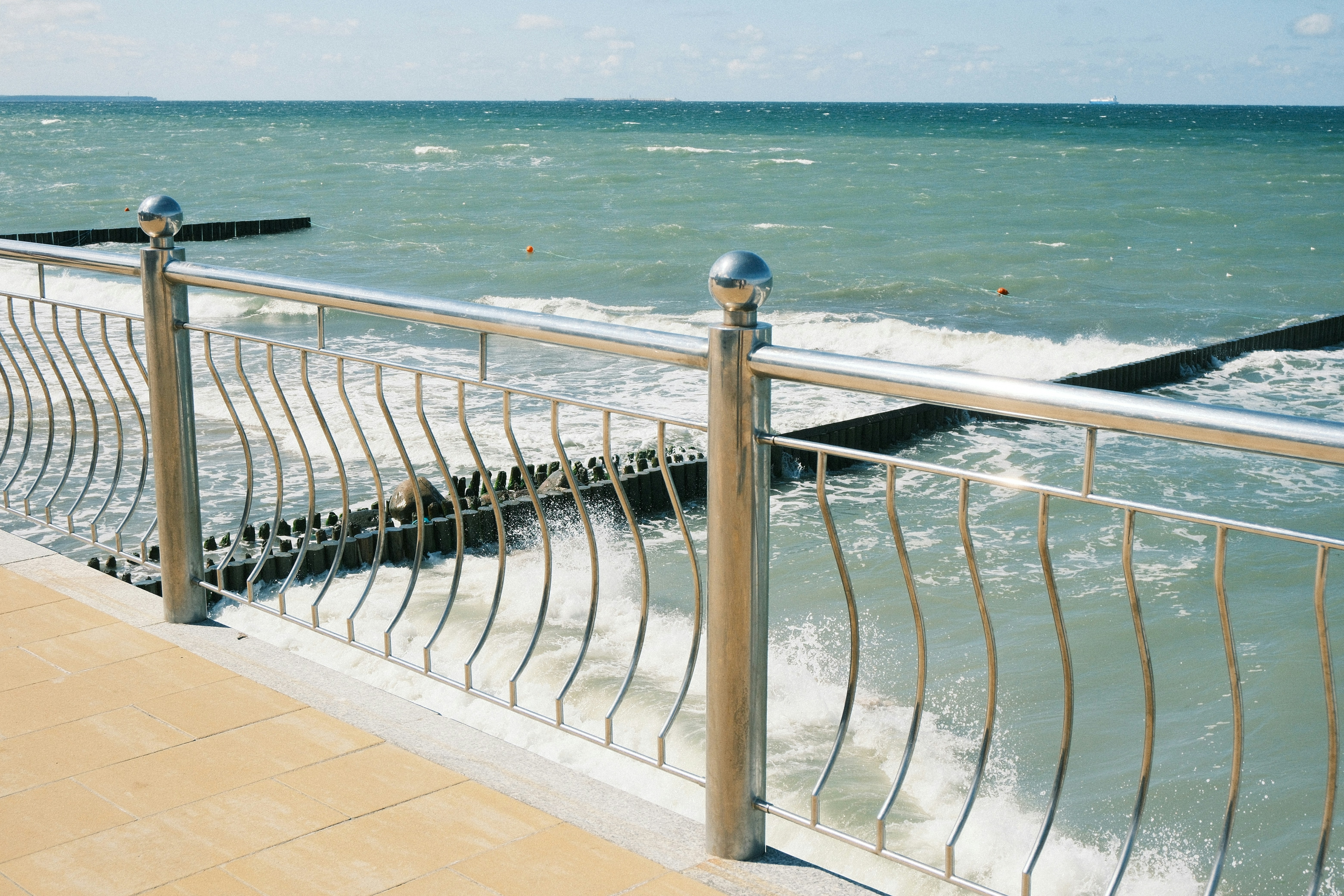 Sunlit seaside railing along a concrete promenade with waves crashing against a barrier under a clear sky.