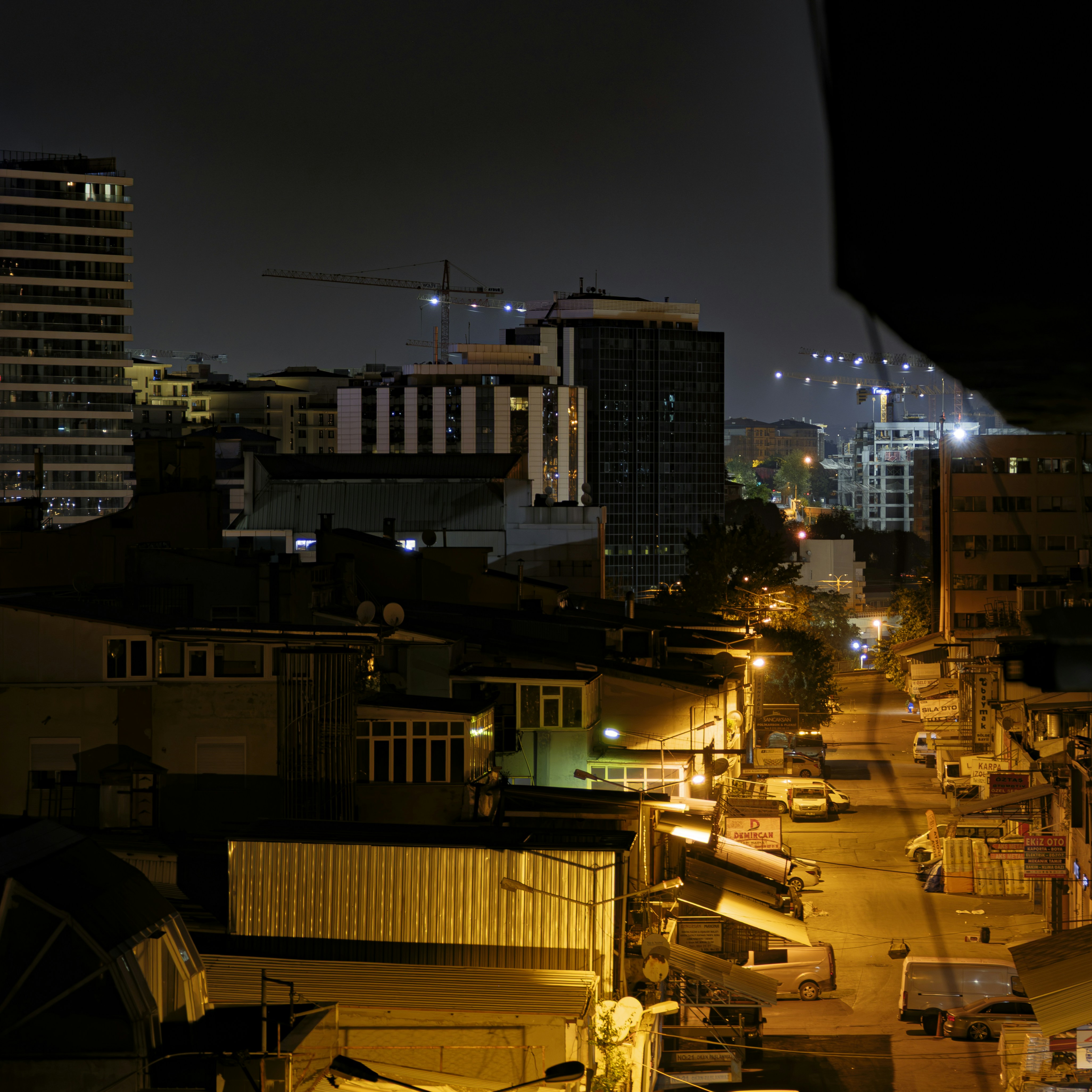 Dimly lit urban street scene with warm streetlights, mid-rise buildings, and distant construction cranes under a dark sky.