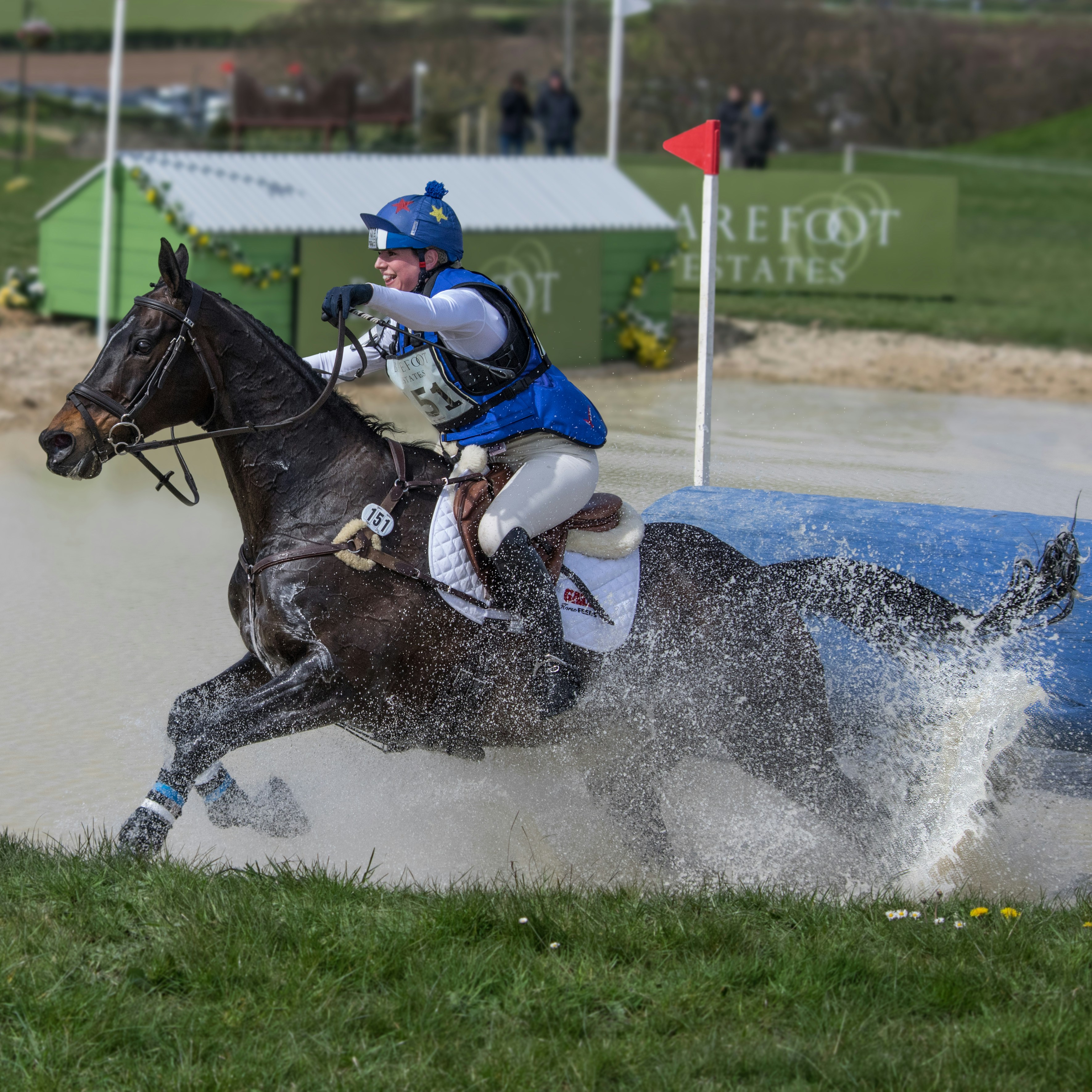 Horse and rider splash through a water obstacle at a cross-country event; the rider leans forward with blue safety vest while the horse powers through spray along a sandy track.