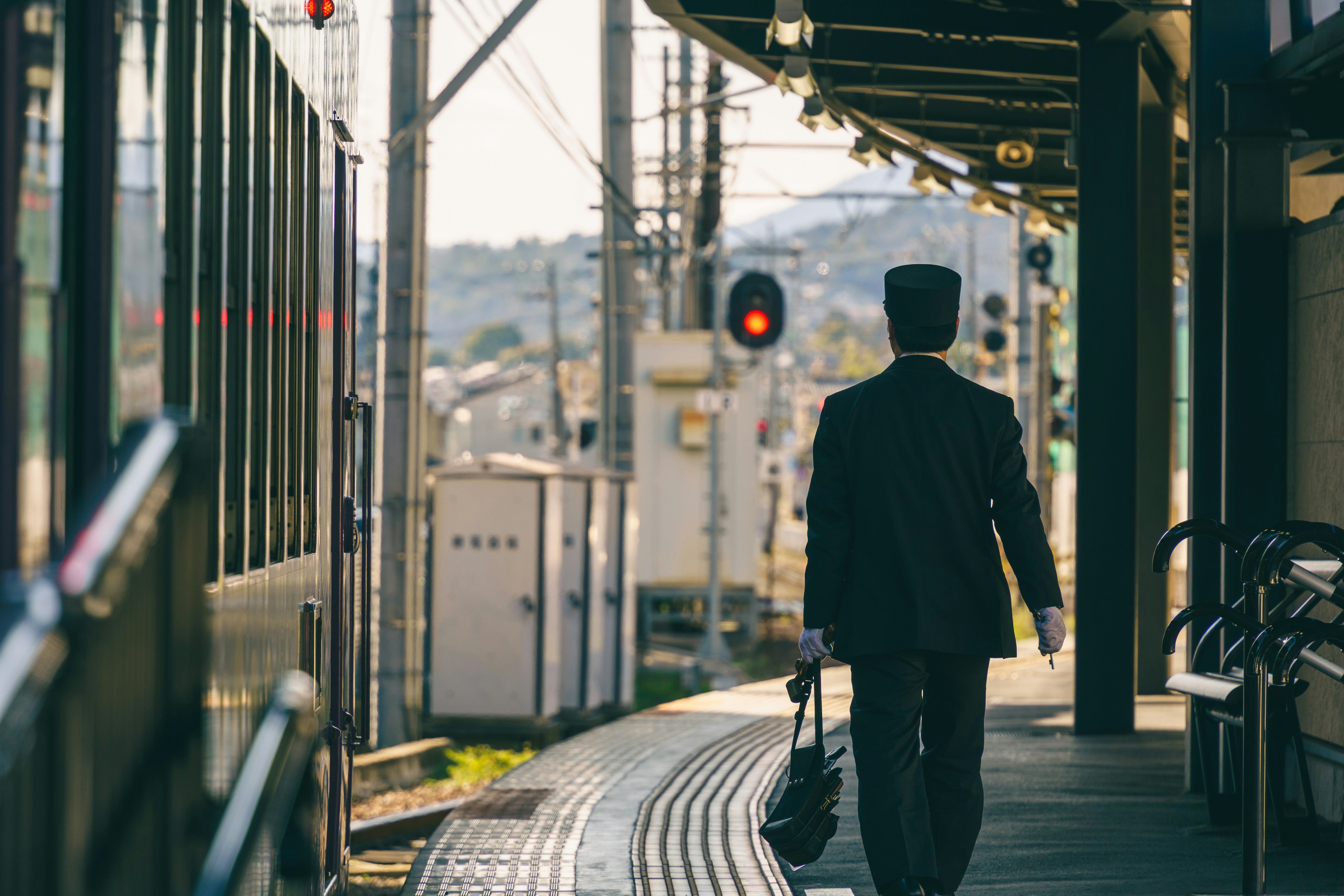 A solitary figure in a dark suit and hat stands on a platform, carrying a bag as trains and rails fade into the distance.