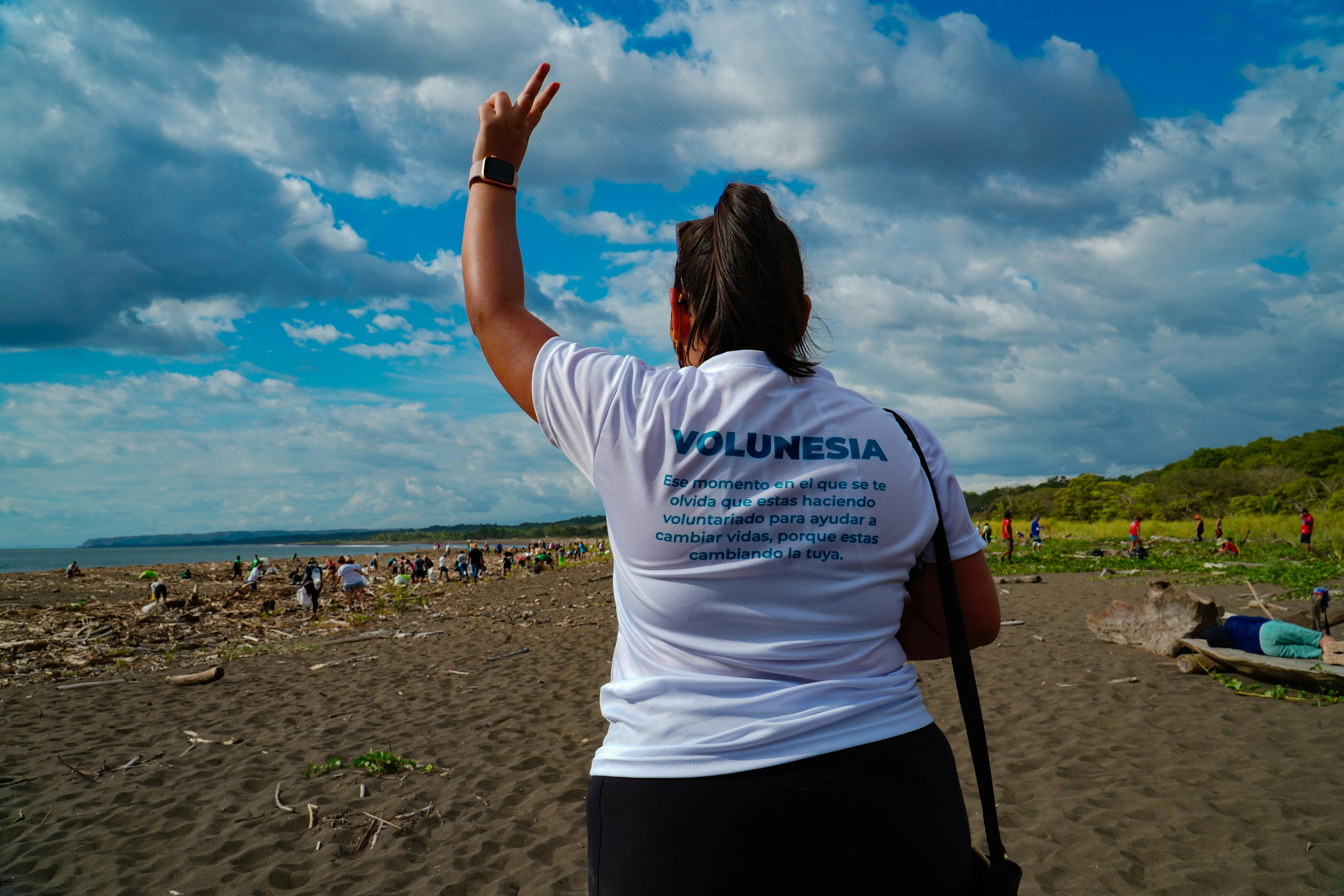 Volunteer wearing a white shirt waves at the horizon on a sandy beach with people and greenery in the background.