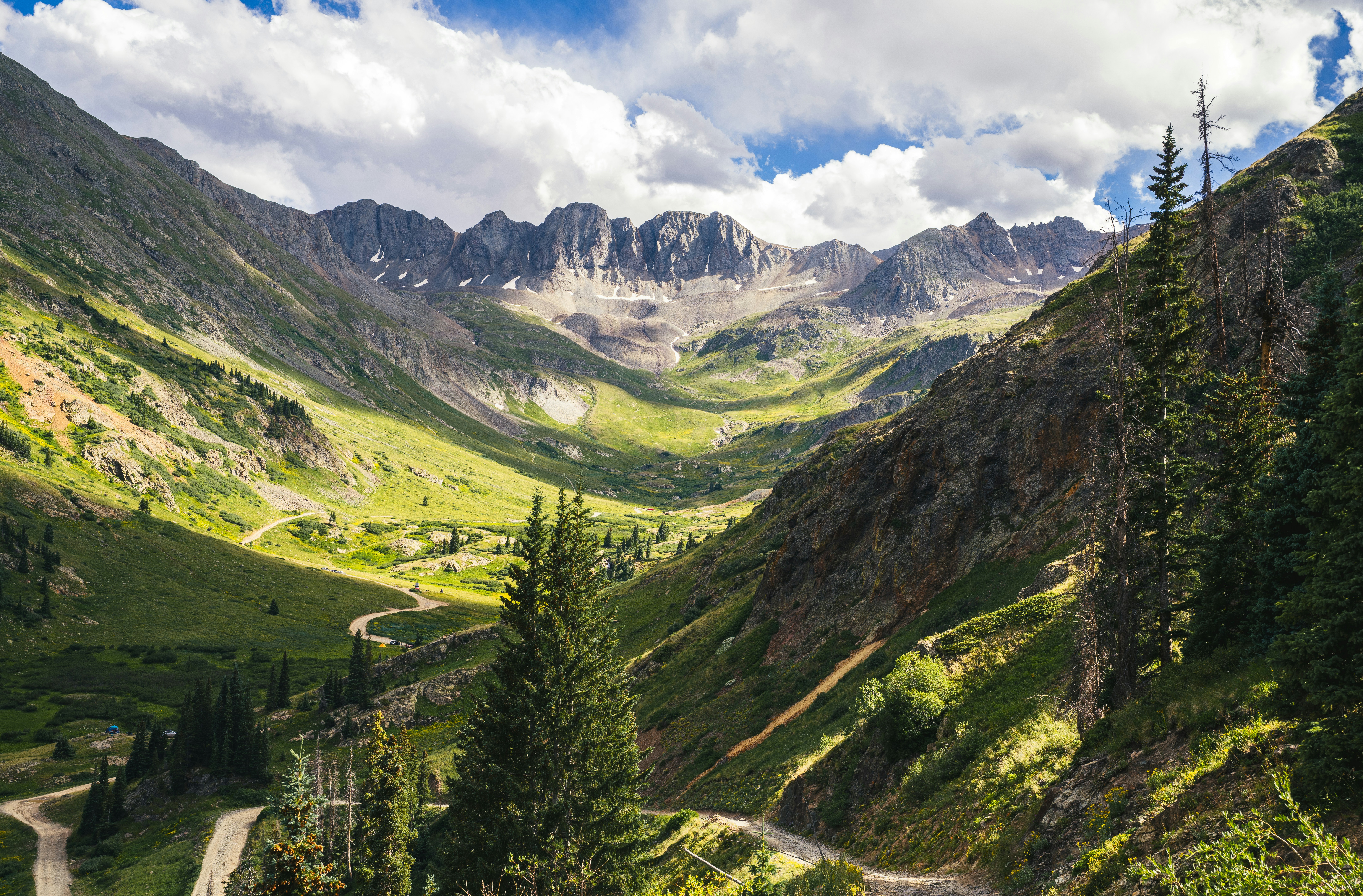 Sunlit alpine valley with winding road and green meadows, framed by rugged peaks and evergreen trees. A landscape scene emphasizing depth and natural color balance.