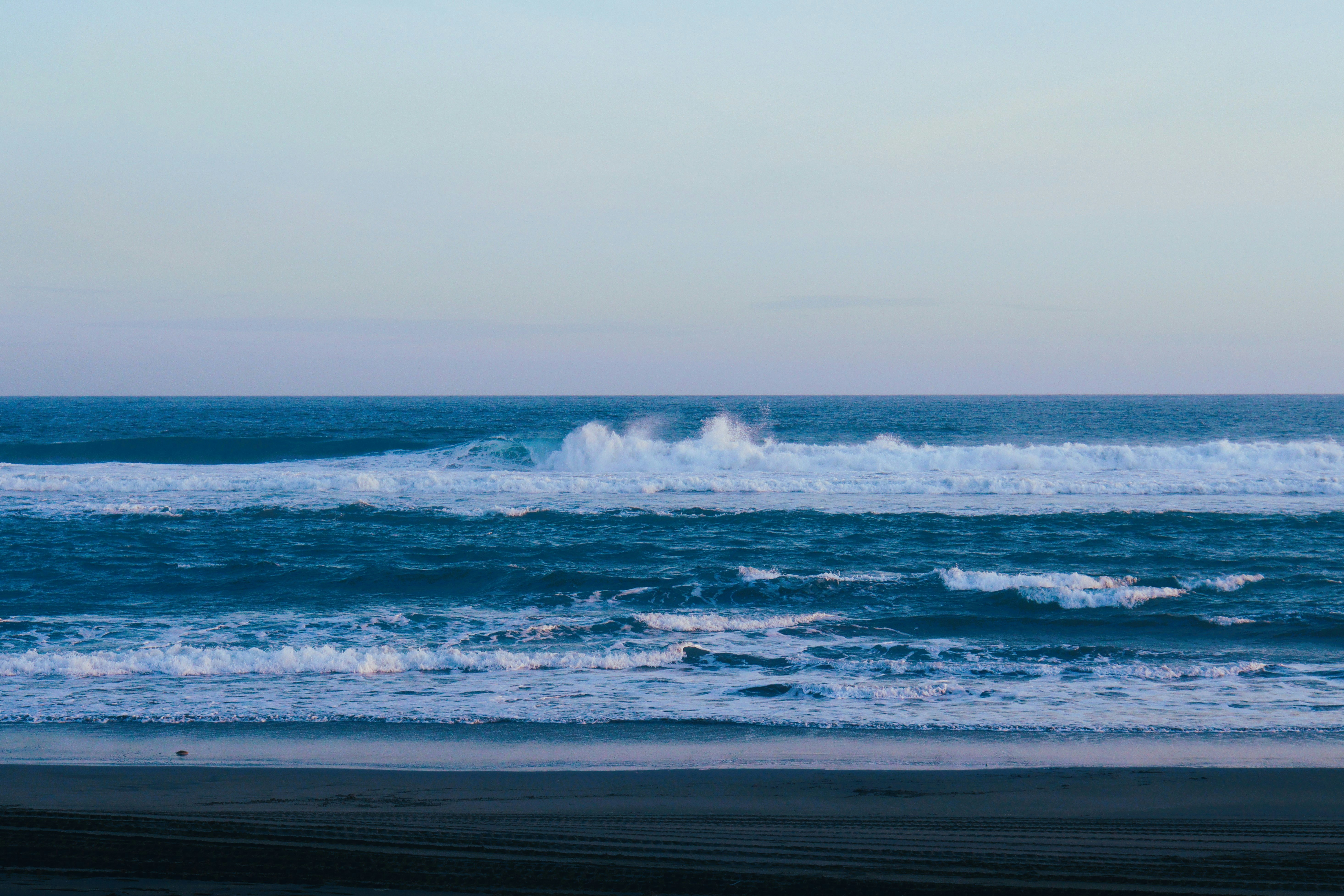 Soft coastal scene with layered waves rolling toward a dark beach under a pale sky.