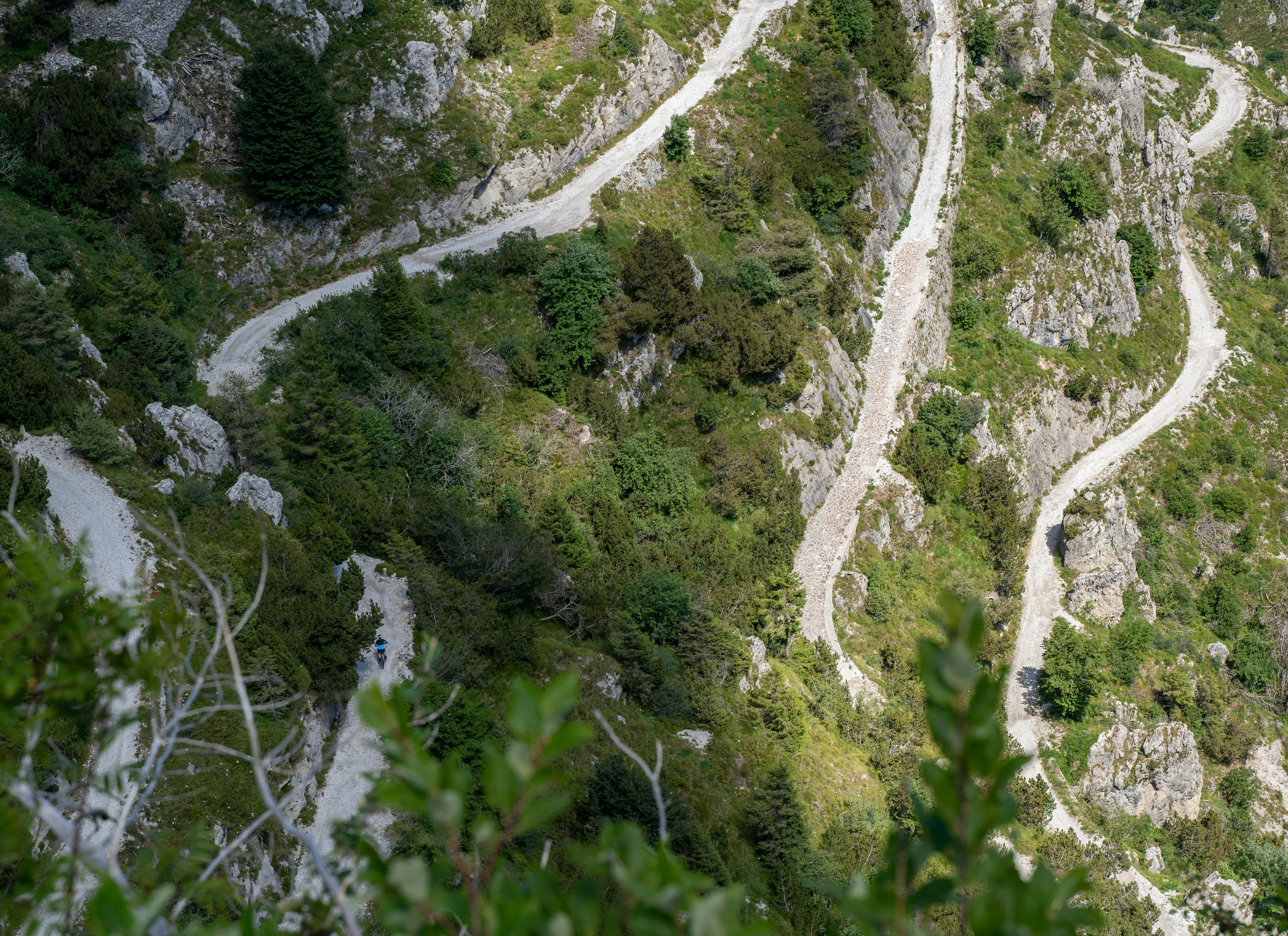 Coiled dirt path threads through rocky, tree-dotted terraced slopes, viewed from above to emphasize the ribbon-like route and varied terrain.