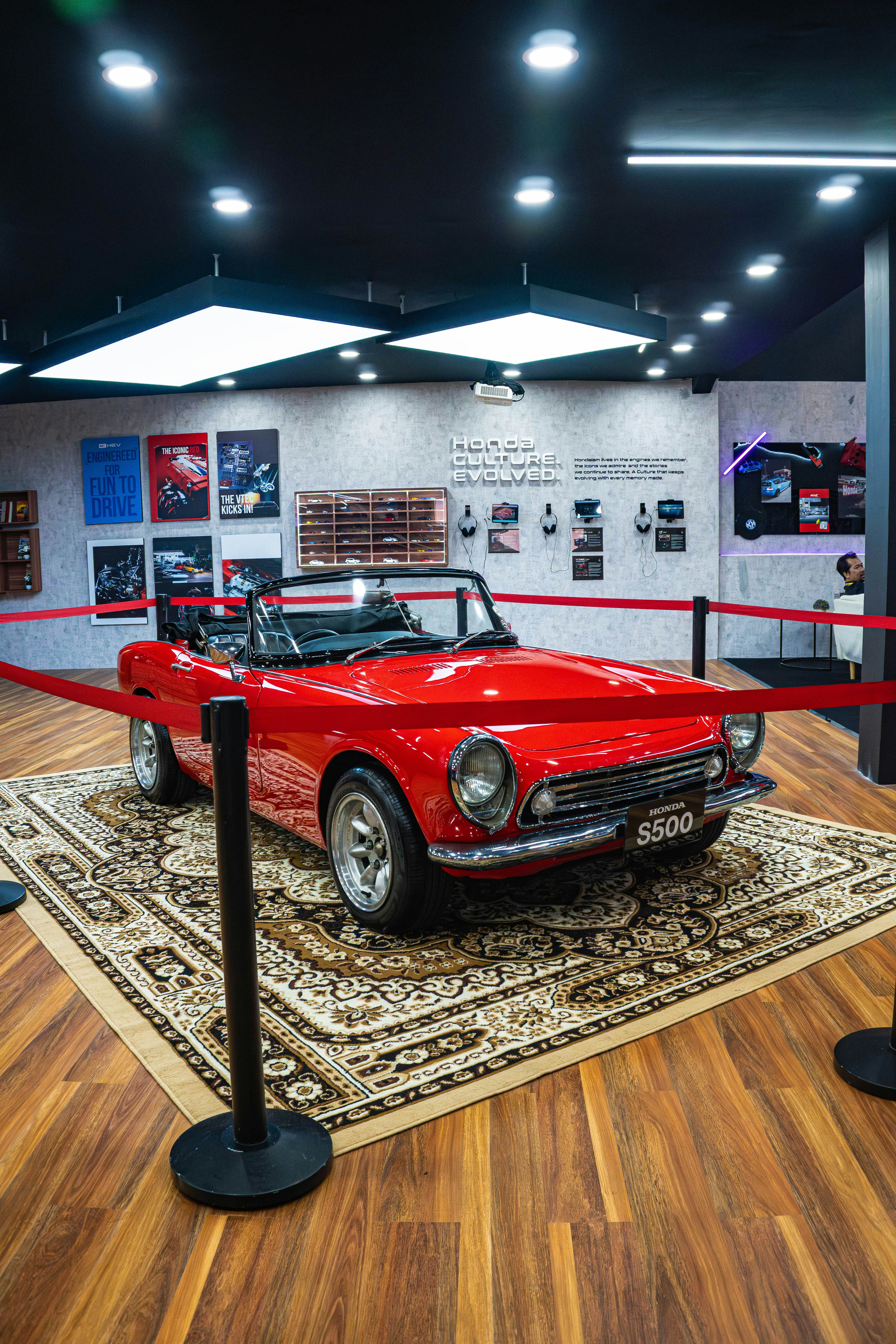Bright red Honda S500 convertible on display in an indoor showroom, roped off with a patterned rug and wooden floor; background features posters and museum-style wall displays.