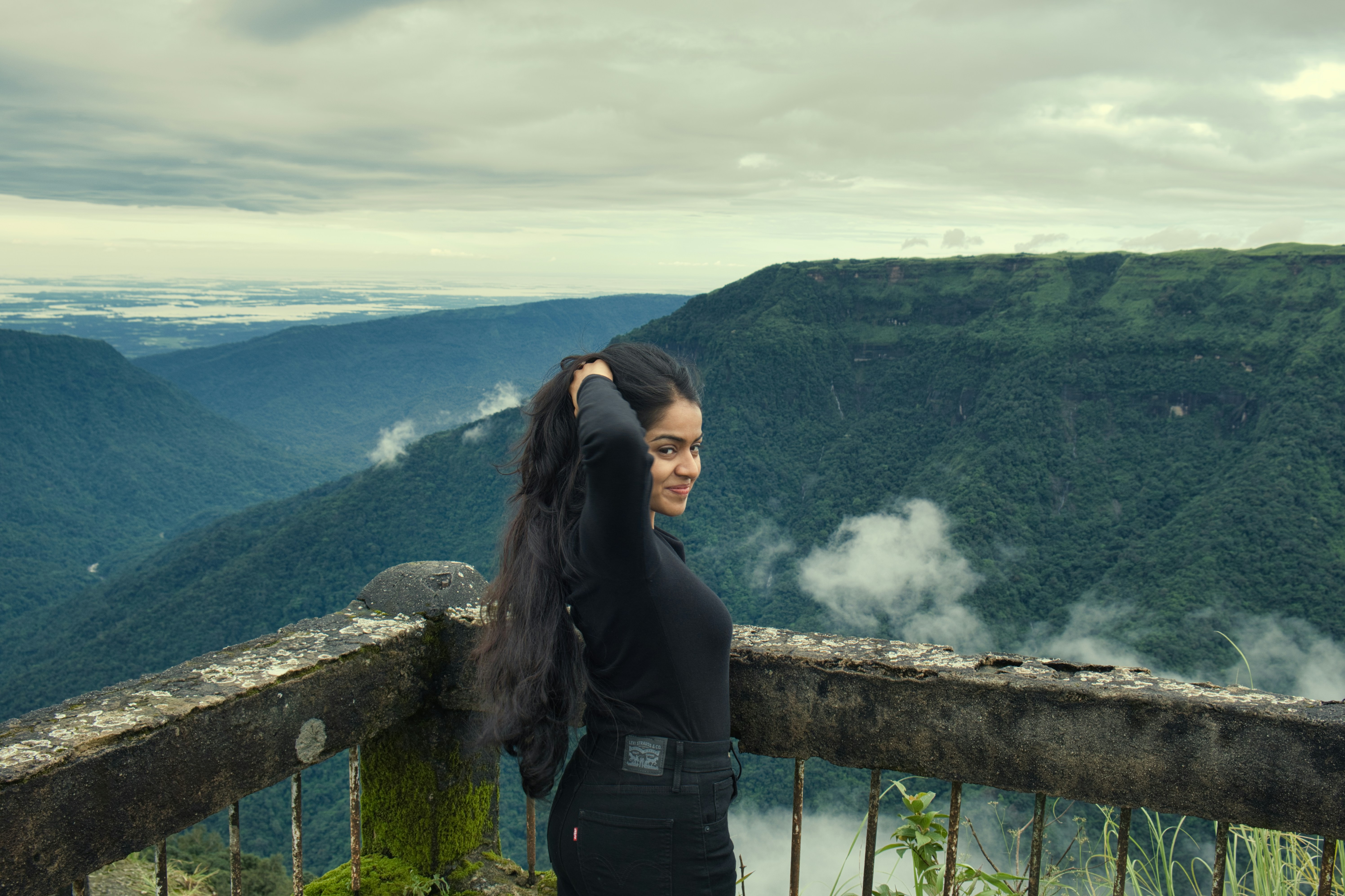Young woman stands at a stone railing on a misty mountain overlook, facing the camera with one hand in her hair; the expansive valley and cloud wisps create a dramatic backdrop. This is a candid portrait with natural scenery.