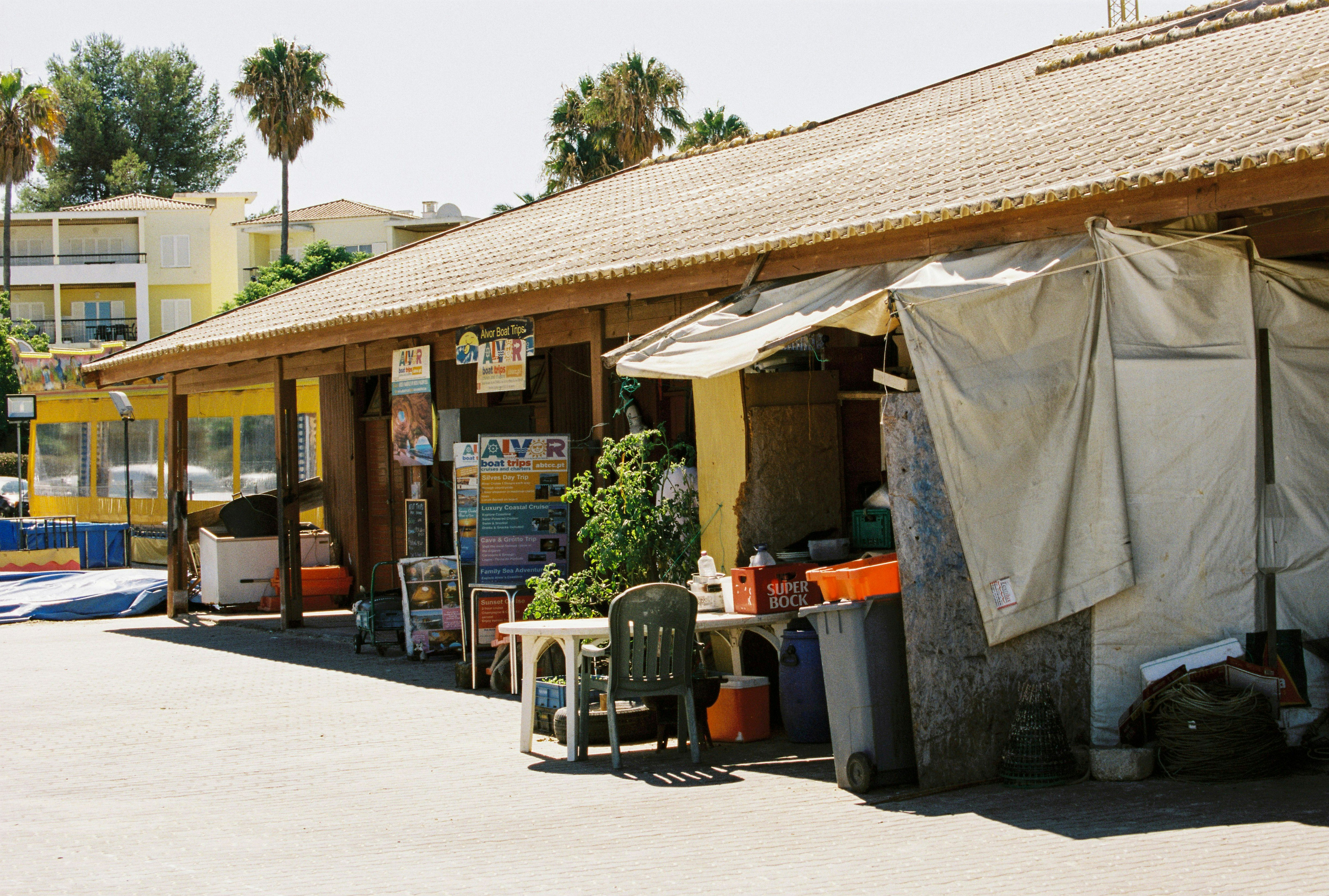 A rustic boat-trip kiosk with palm trees and colourful signage advertises Algarve day trips under a tiled roof.