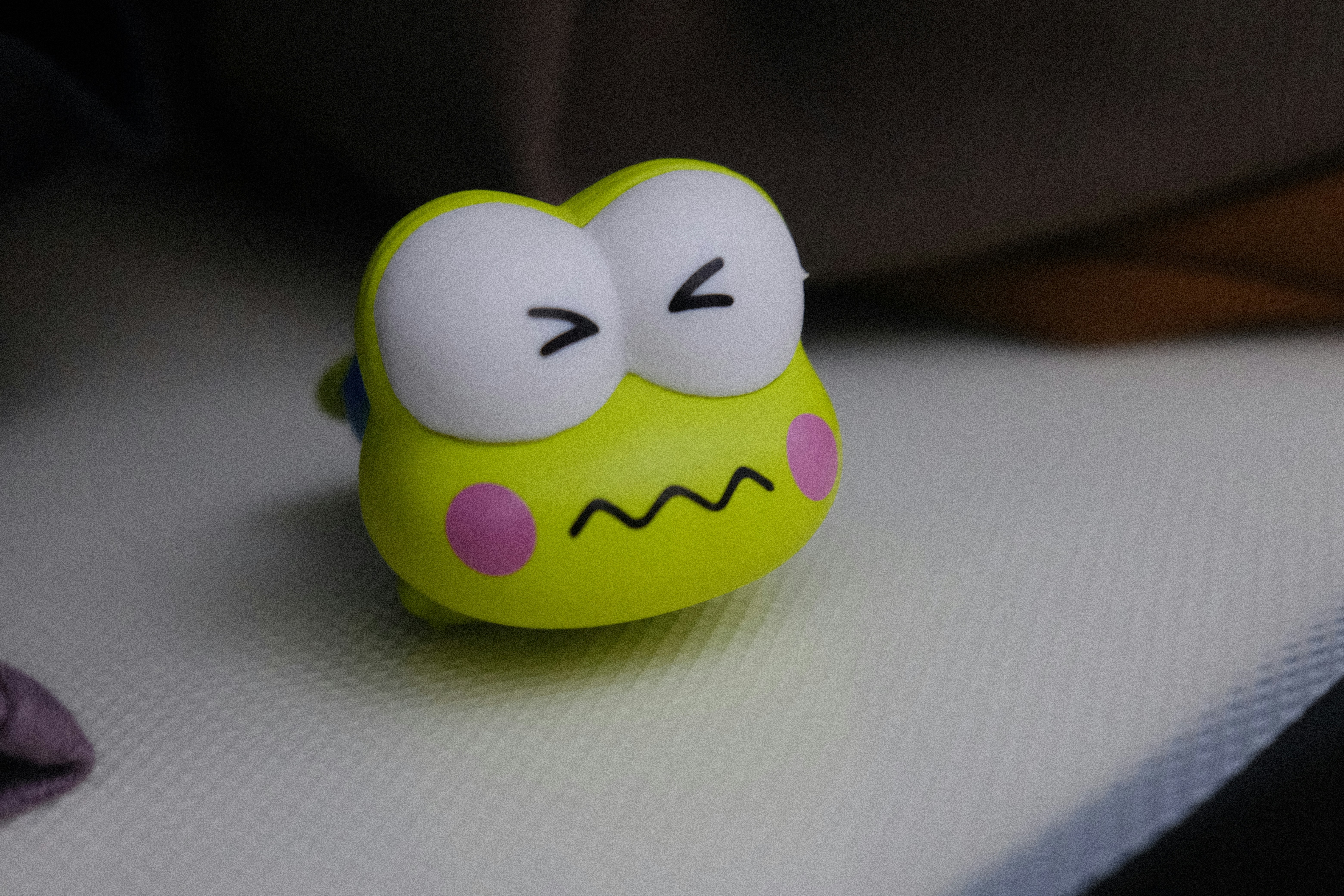 Bright yellow cartoon squishy with pink cheeks and squinting eyes sits on a textured white surface, softly lit from the side.
