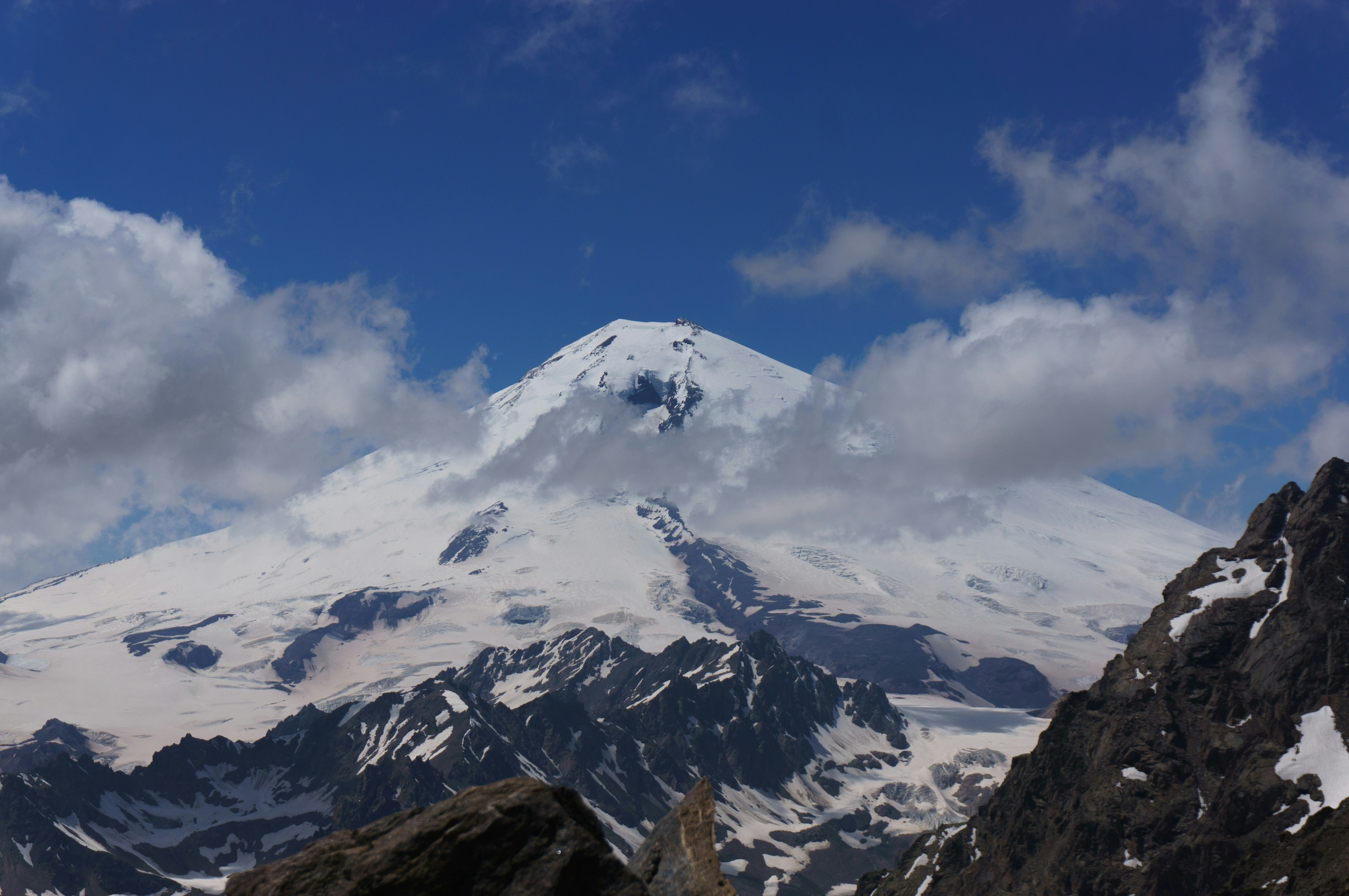 Snow-capped summit rising above rugged foothills with clouds drifting around, set against a blue sky.
