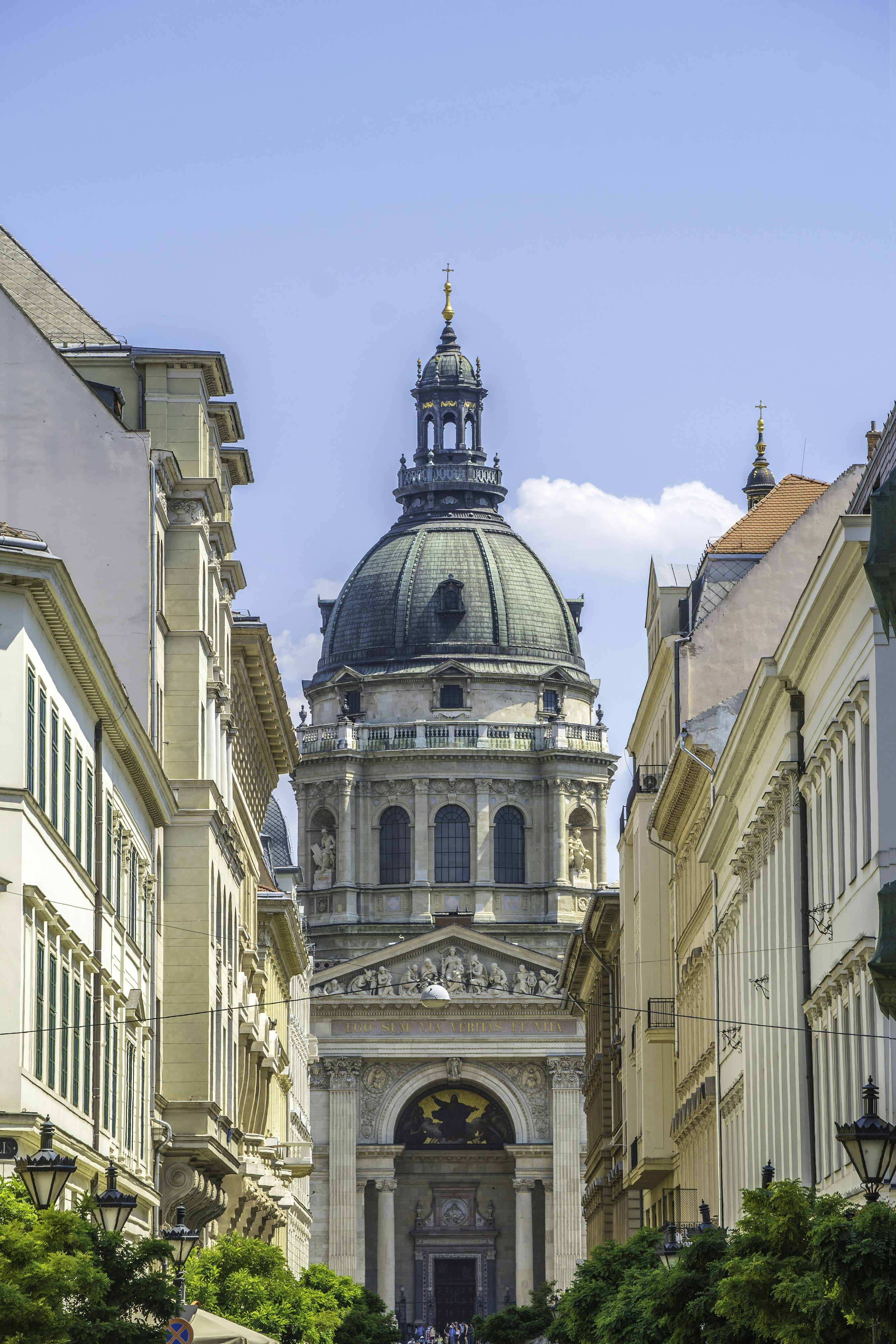 Stone cathedral with a dominant copper-green dome rises between lined city buildings; a narrow street leads the eye toward the ornate façade and central arch. Photograph captures urban framing around a religious monument.