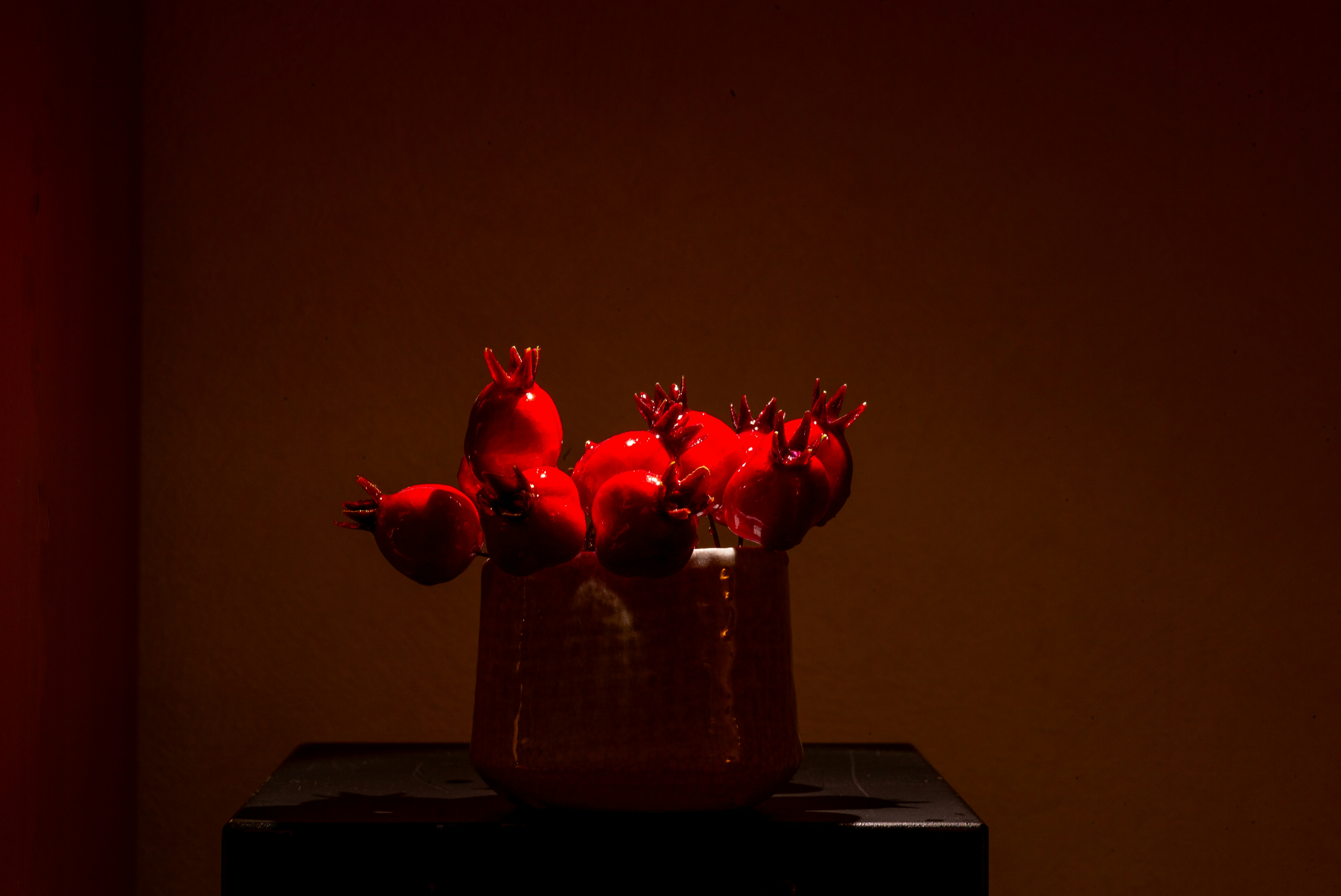 A small clay vase holding bright red pomegranate fruits sits on a dark pedestal, illuminated by a warm, moody light.