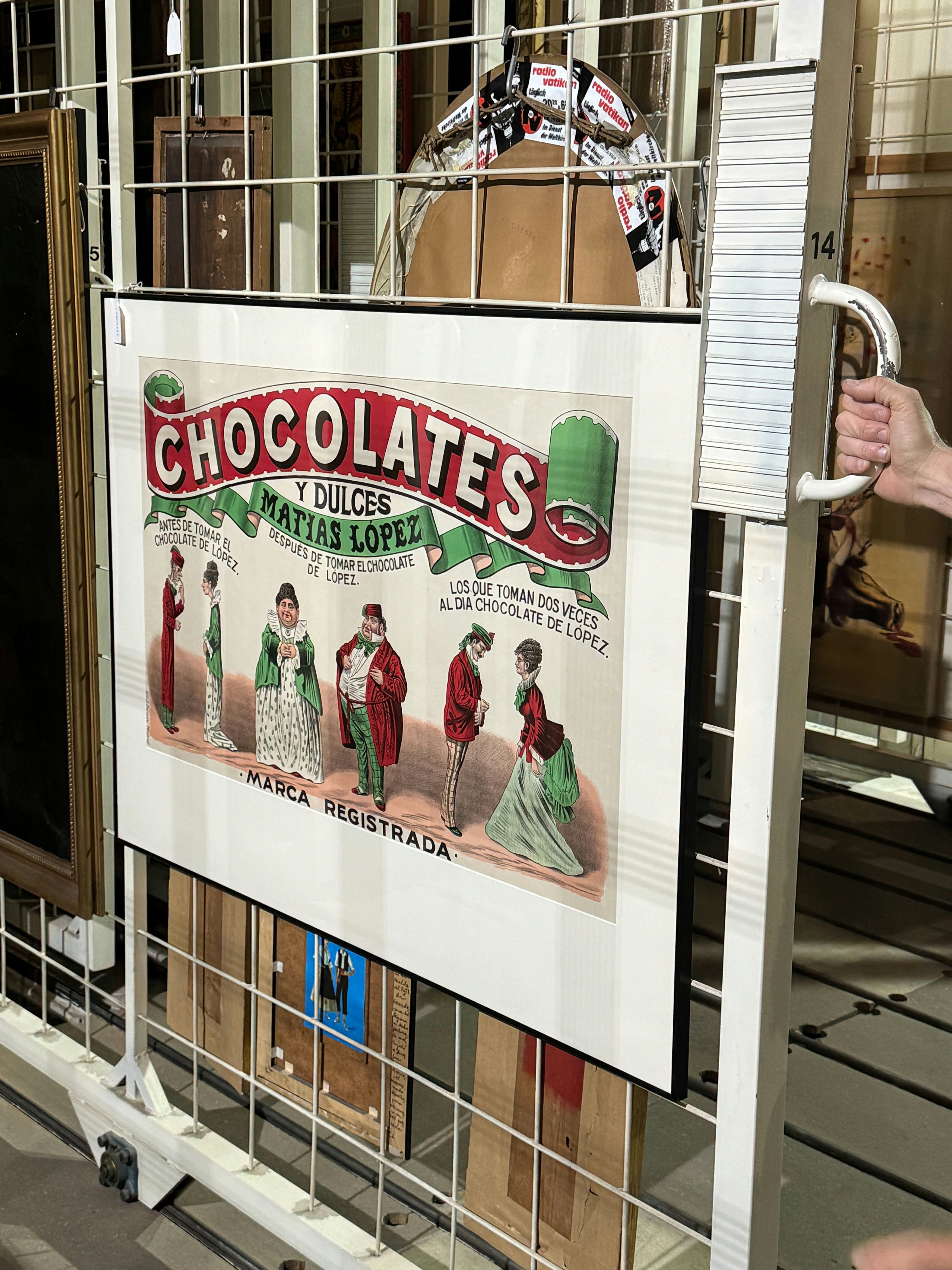 Poster for chocolates and Dulces Lopez secured in a wheeled metal rack; the frame shows a historic advertising illustration with people in period attire. Acrylic-print poster in a retail or exhibit setting.