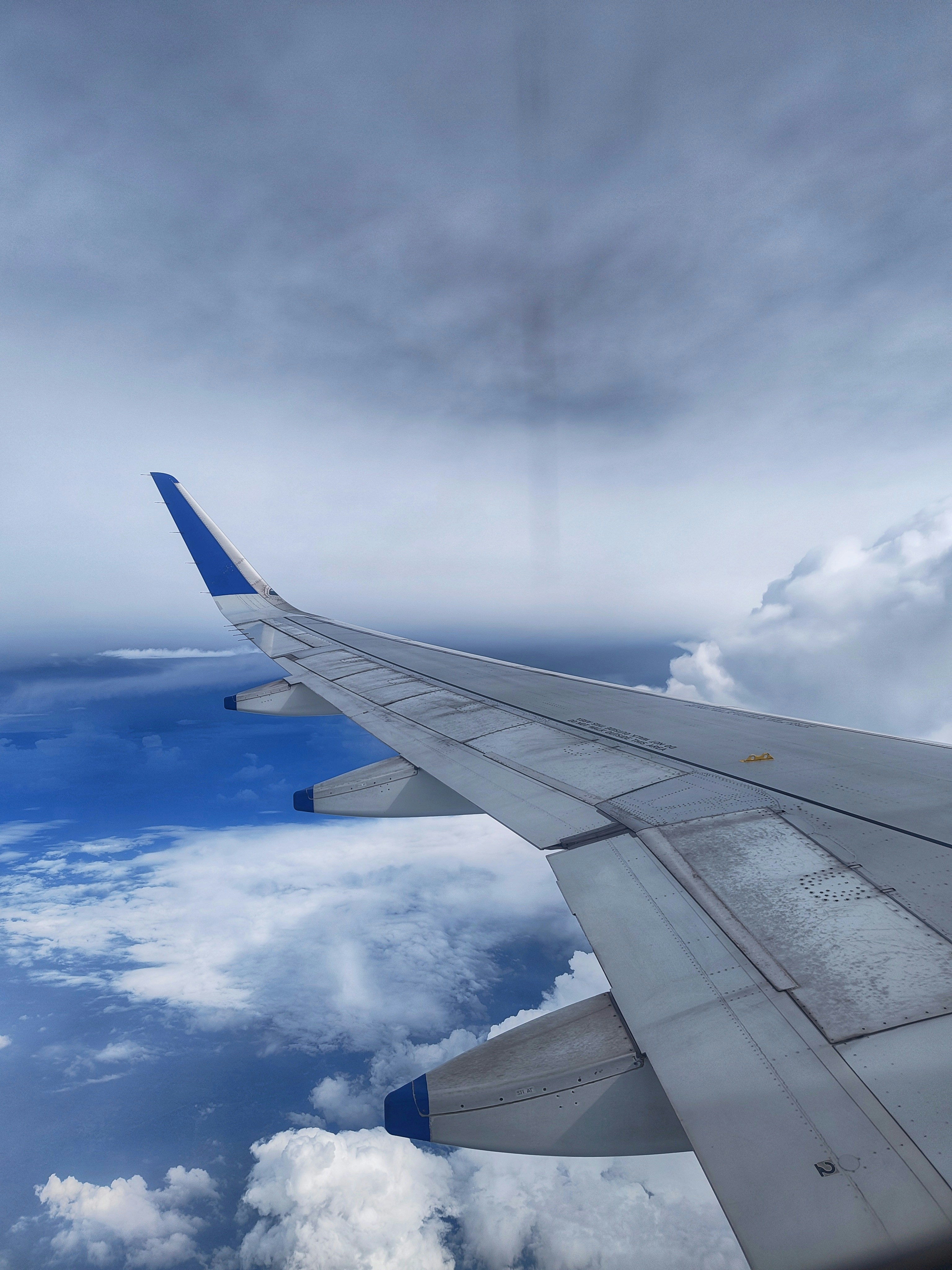 Airplane wing cuts through a bright blue sky with wispy clouds below, creating a dramatic aerial view from the window. The scene captures altitude and perspective; the image emphasizes the aircraft structure and expansive horizon.