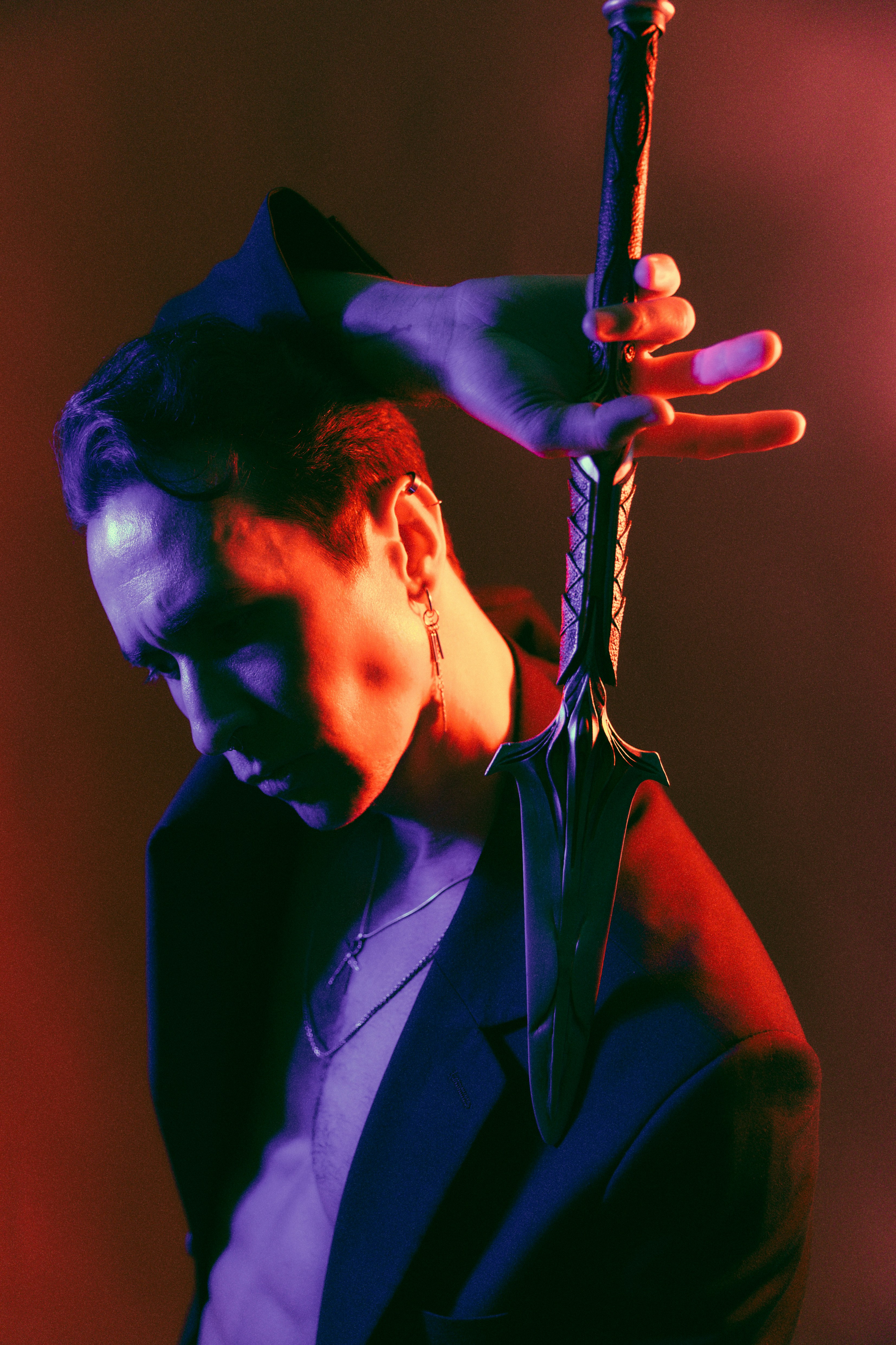 A moody portrait of a dancer in a dark suit, head bowed, gripping a decorative blade against a red-blue backlit background. This is a photography still, not a painting or logo.