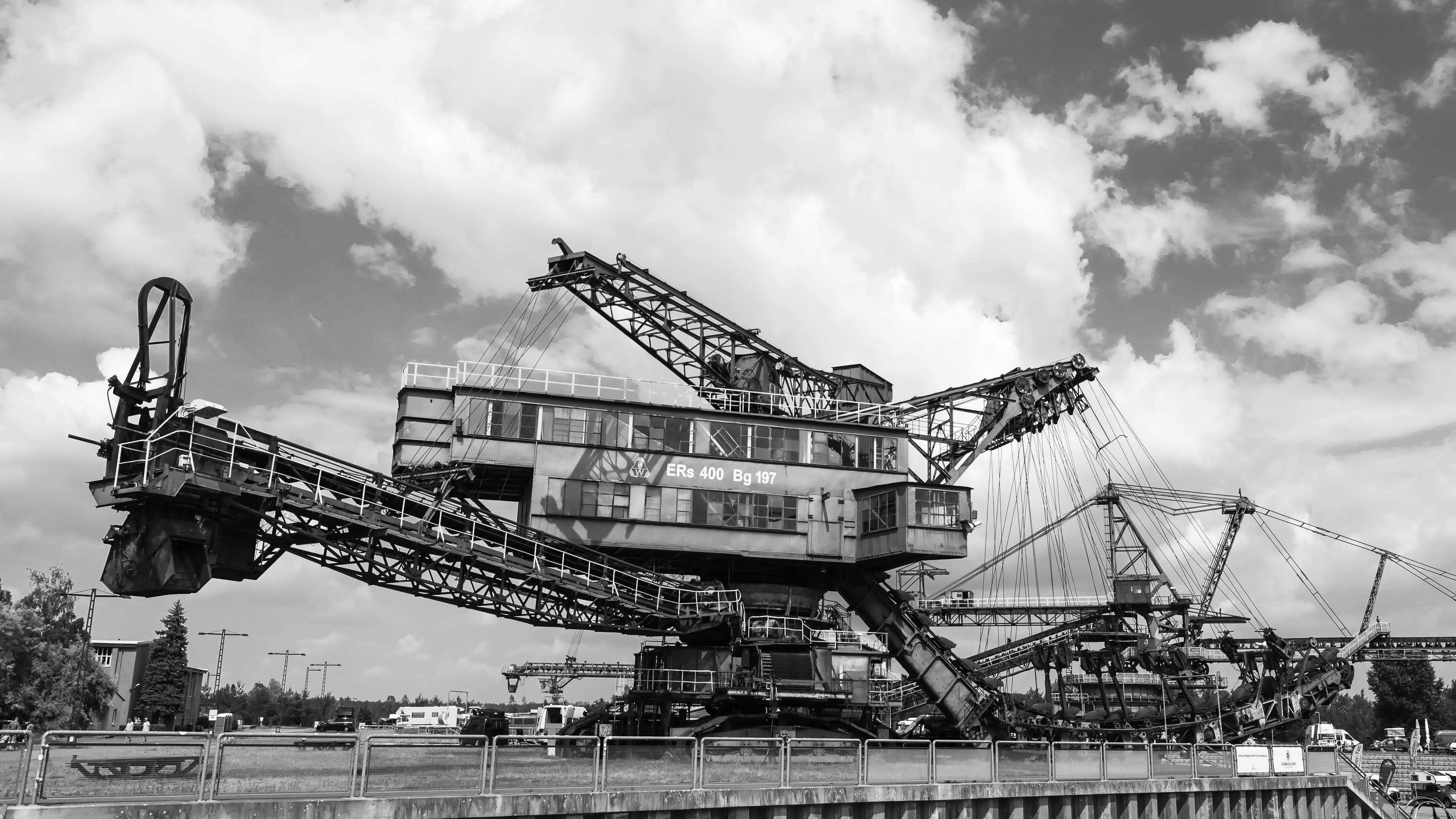 Monochrome scene of a colossal mining crane dominating a flat landscape, with dramatic clouds and distant vehicles providing scale.