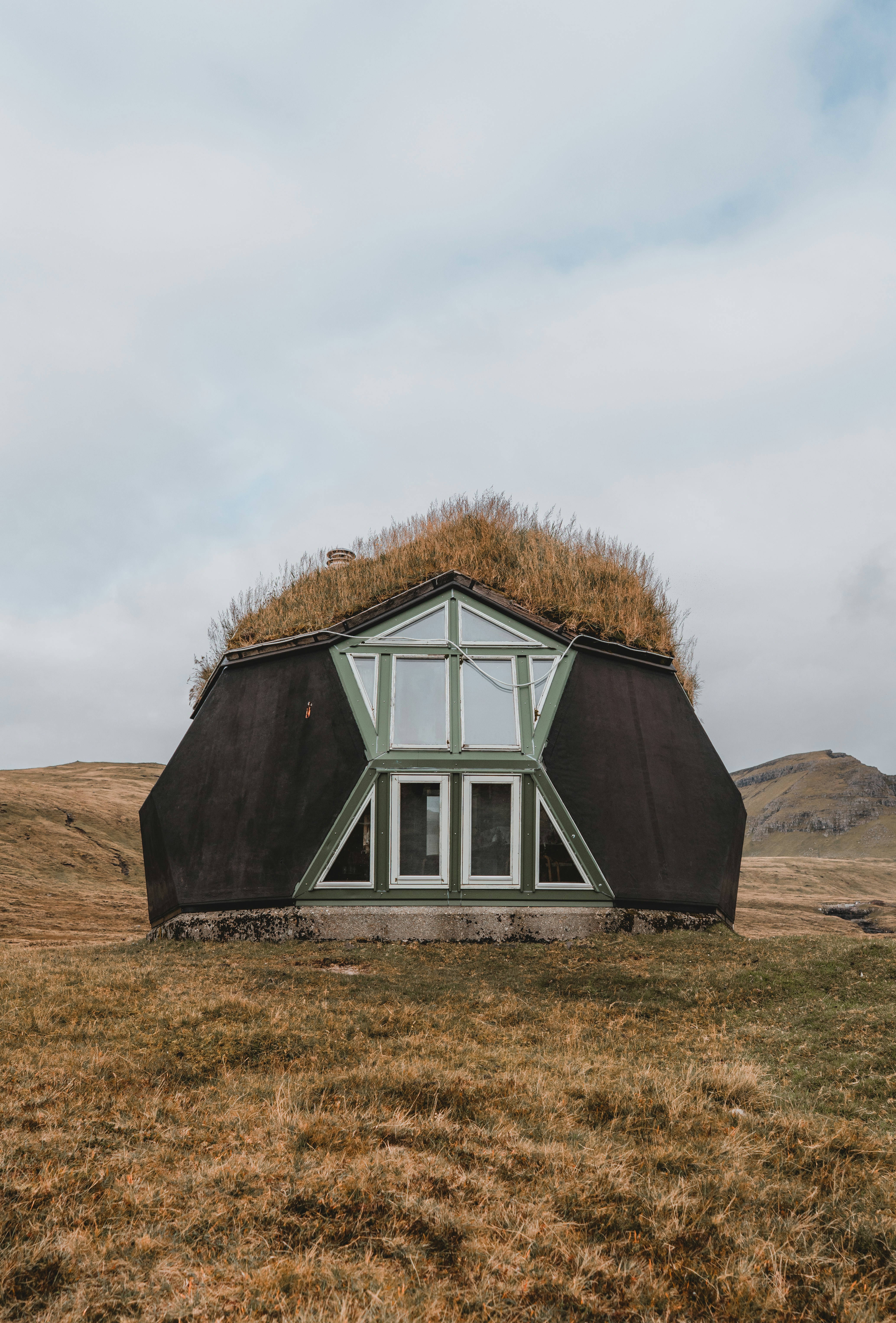 A small geodesic dwelling with grass-covered roof sits on a windswept plain beneath a pale sky, its triangular windows framing the landscape.