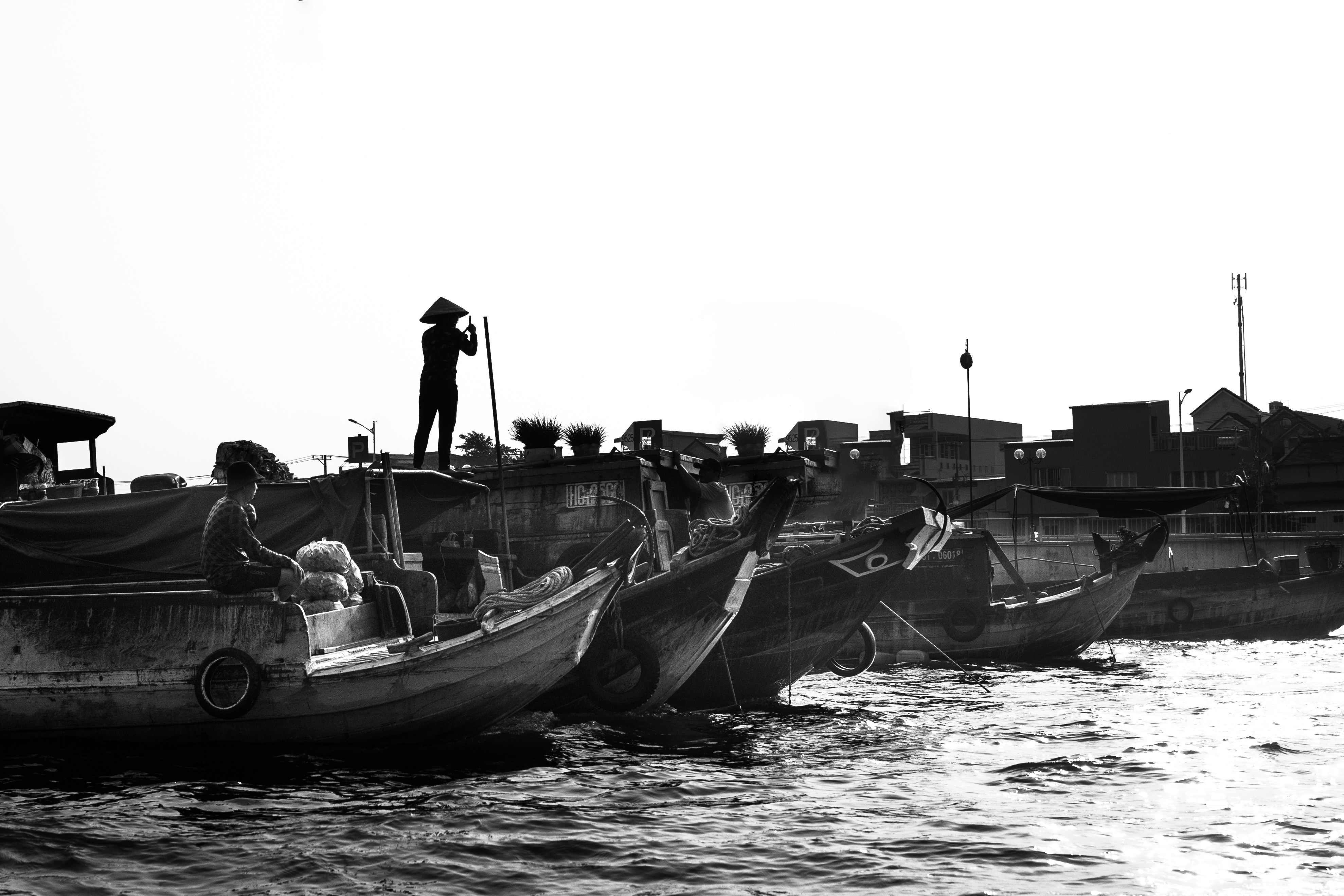 Black-and-white scene of a harbor with multiple boats docked along the quay; a tall figure stands on one boat, perhaps a fisherman, against a bright sky. The image emphasizes silhouettes and water reflections.