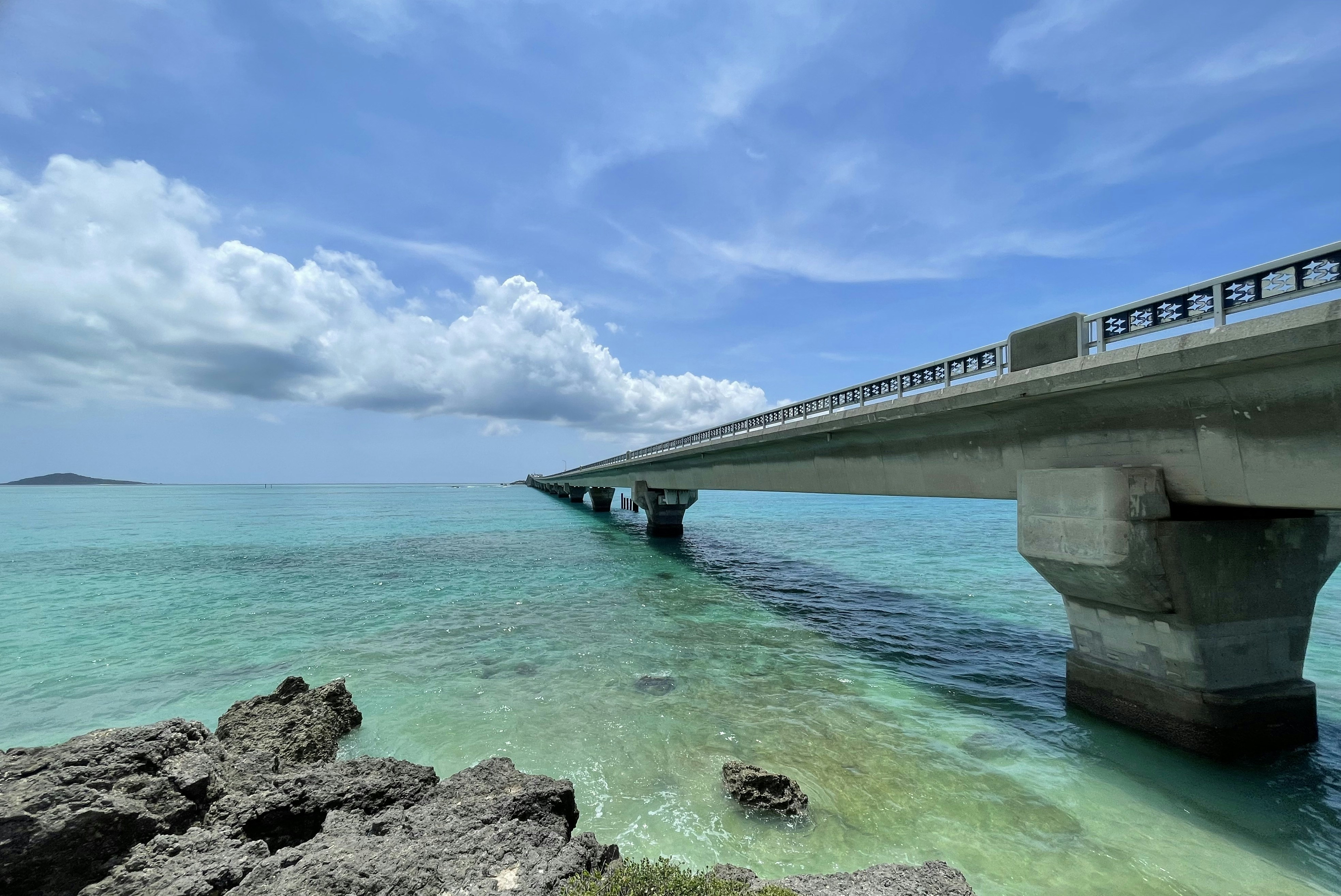 Sunlit coastal scene with a concrete bridge stretching over clear turquoise waters toward the horizon, rocks in the foreground and a blue sky with scattered clouds.