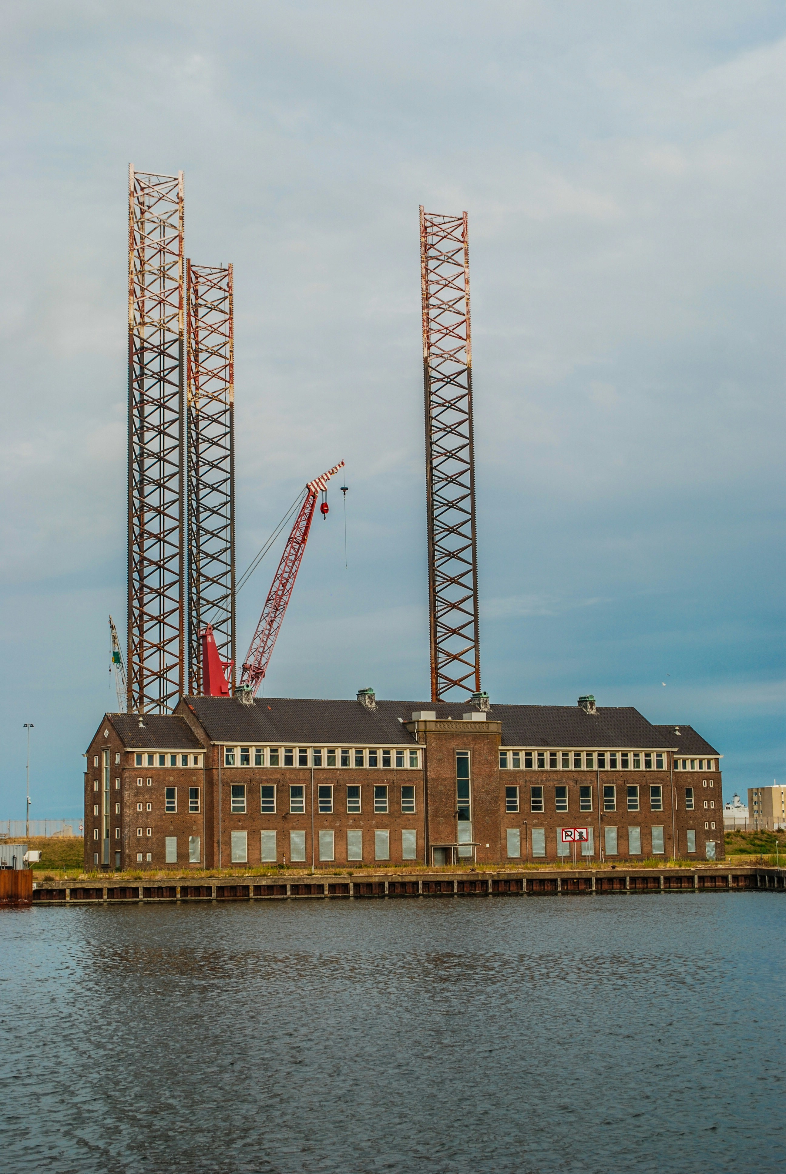 Brick warehouse overlooking calm water with tall structural frames and a crane at the harbor edge.